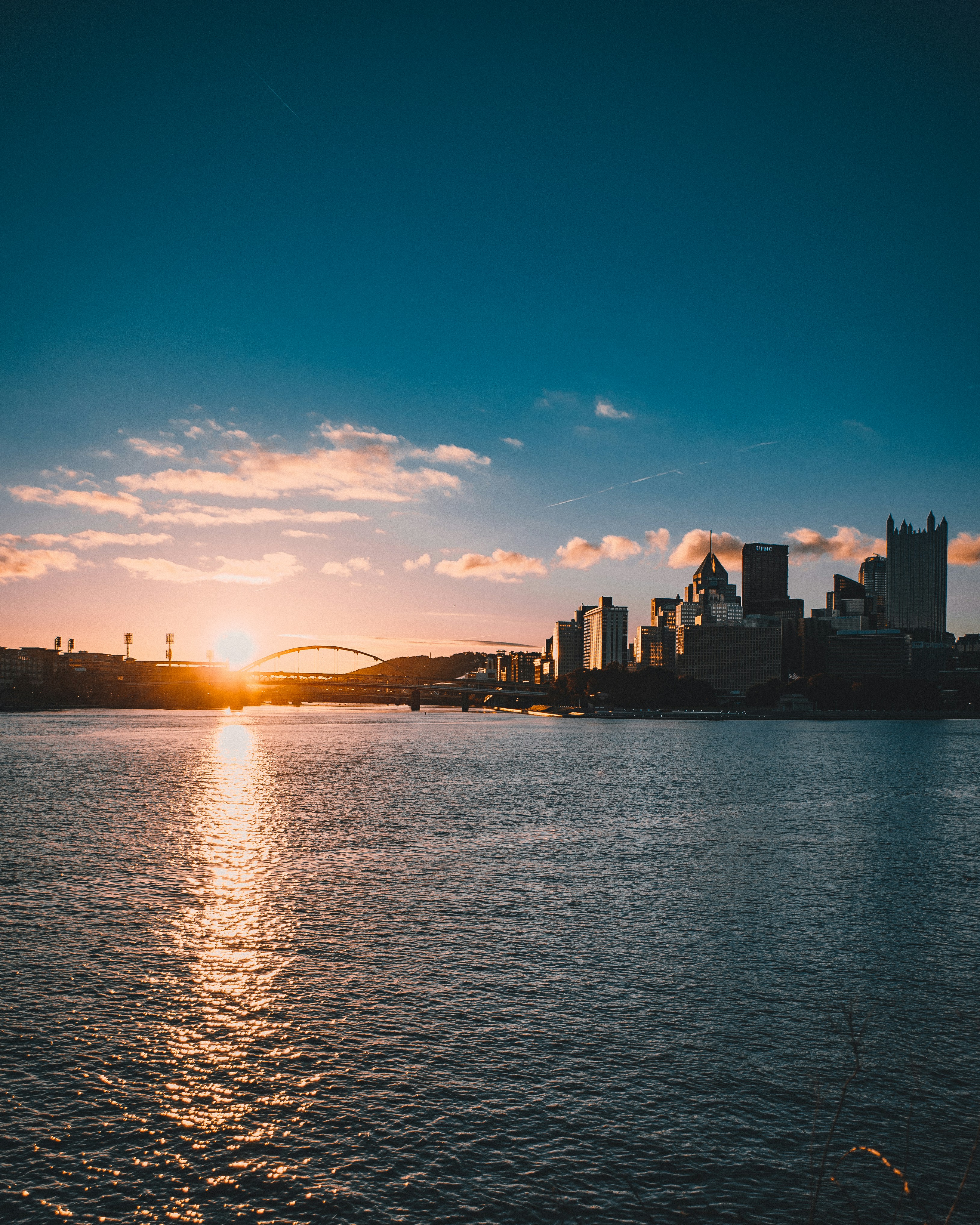 Sunset casting golden hues over a city skyline, with a bridge arching over the water. The calm river reflects the vibrant colors of dusk.