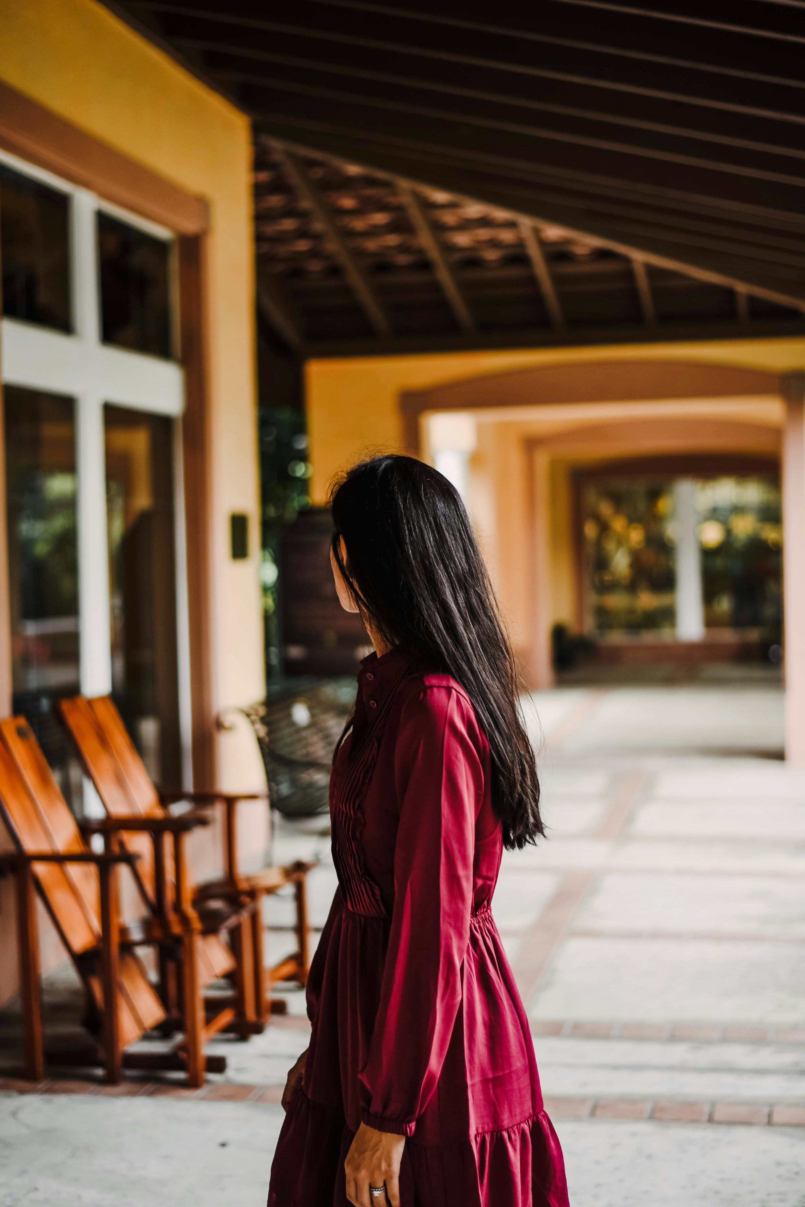 A woman in a flowing burgundy dress gazes thoughtfully towards a tranquil walkway adorned with wooden chairs and soft lighting.