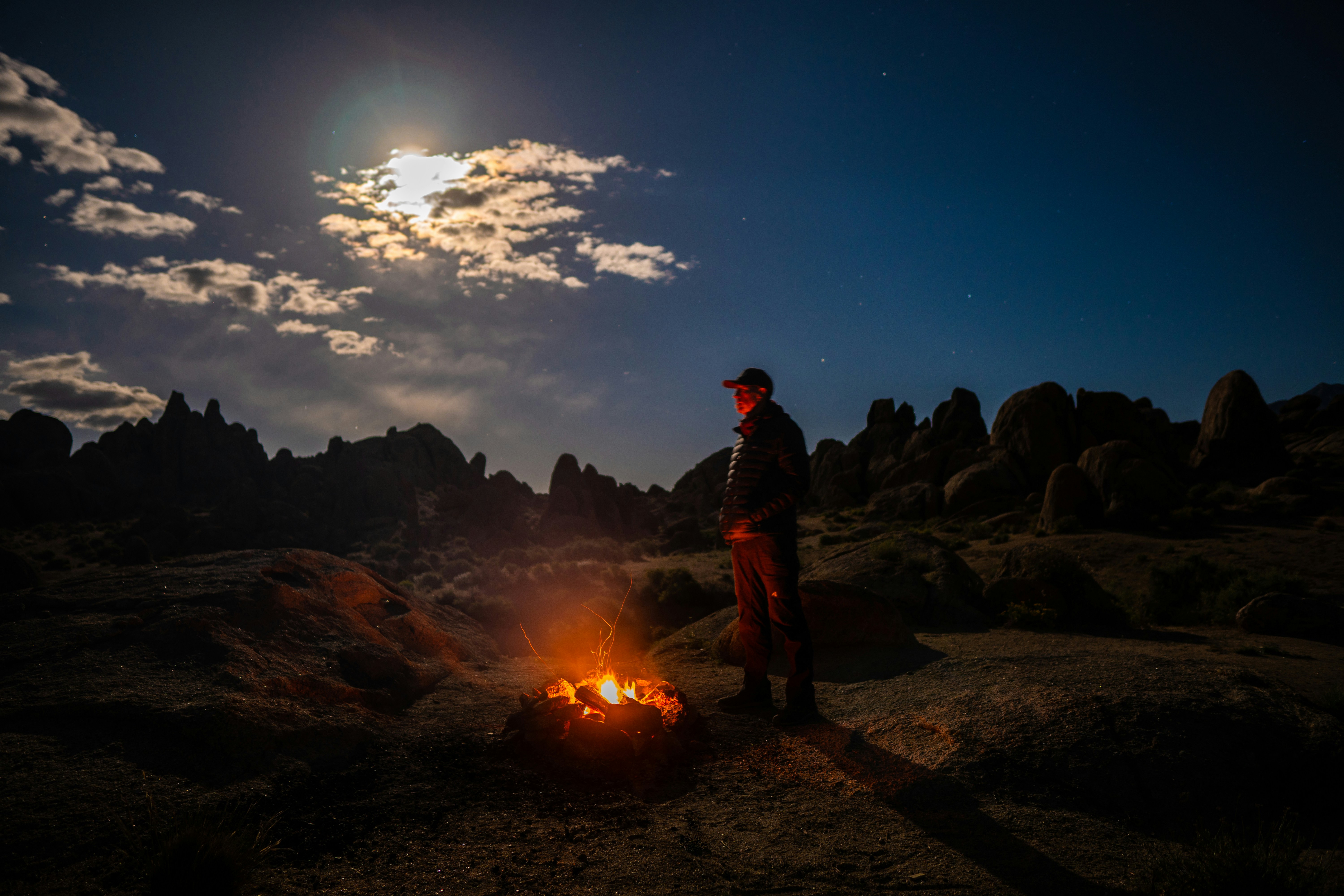 A figure stands beside a glowing campfire under a bright moon, surrounded by rugged rock formations. The scene captures the tranquility of a night in nature.