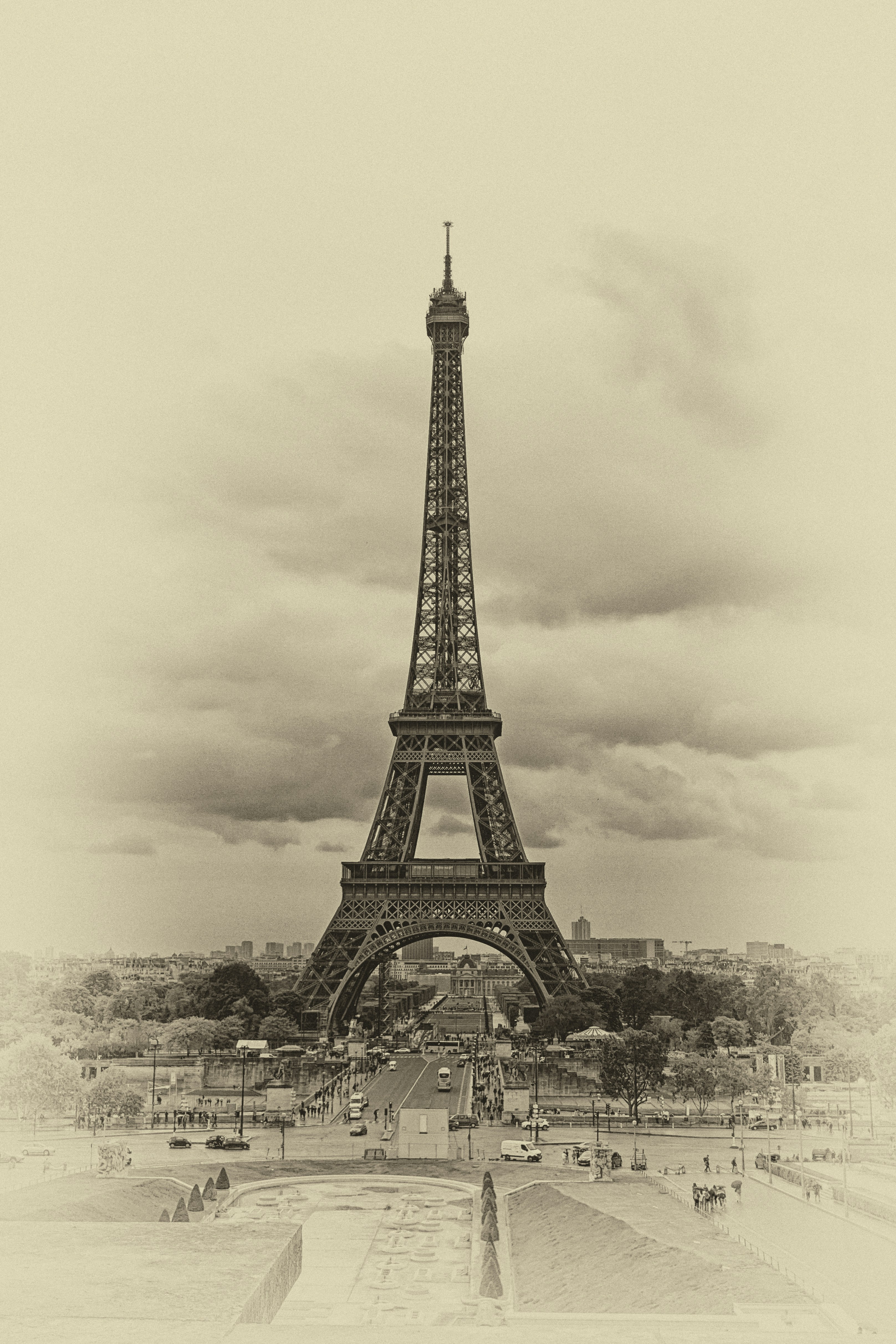 Eiffel Tower rising majestically against a cloudy sky, captured in a sepia tone that adds a vintage charm. The surrounding landscape features pathways and greenery.