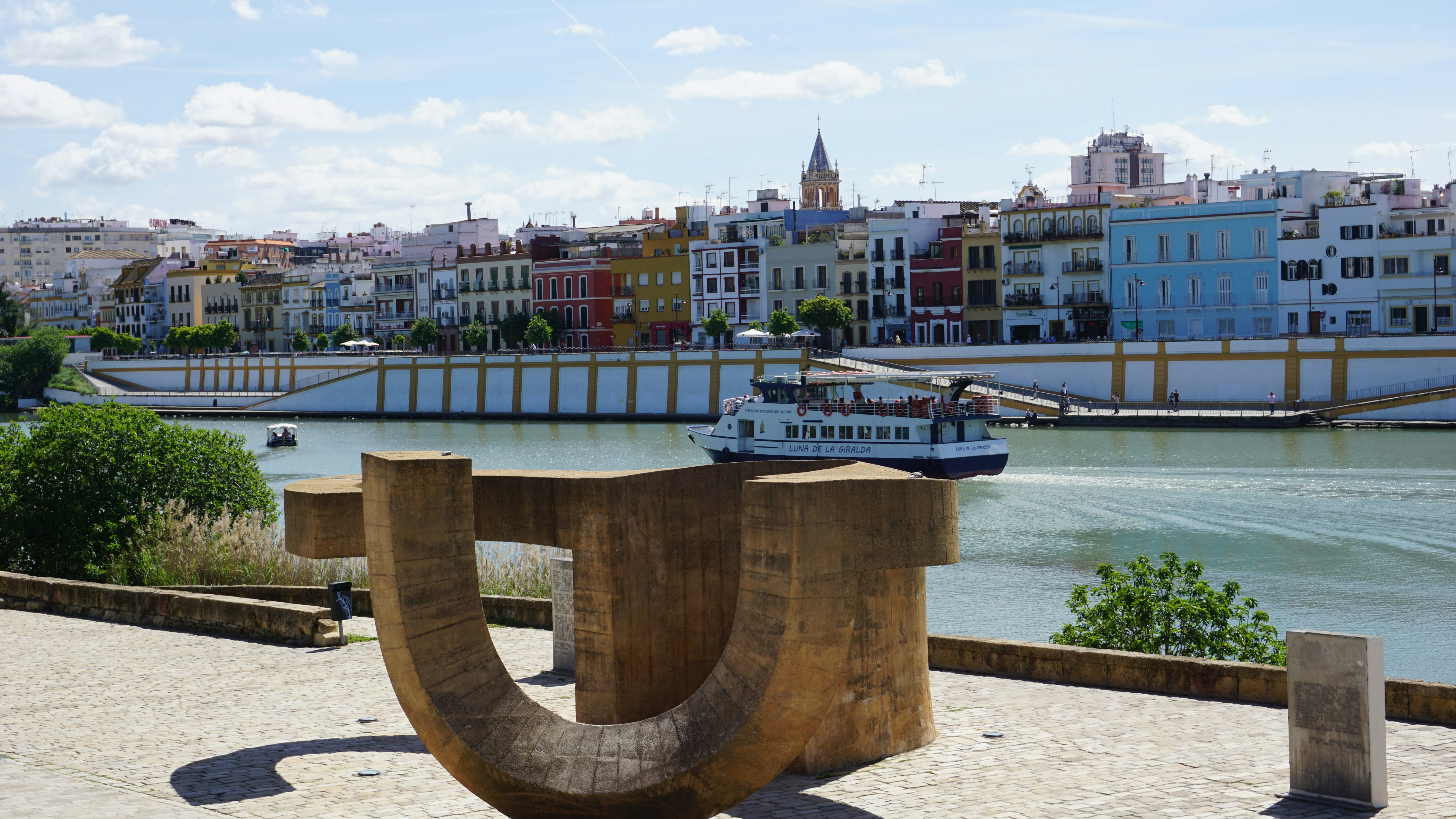 Colorful buildings line the banks of the Guadalquivir River under a sunny sky, with a boat navigating the calm waters.