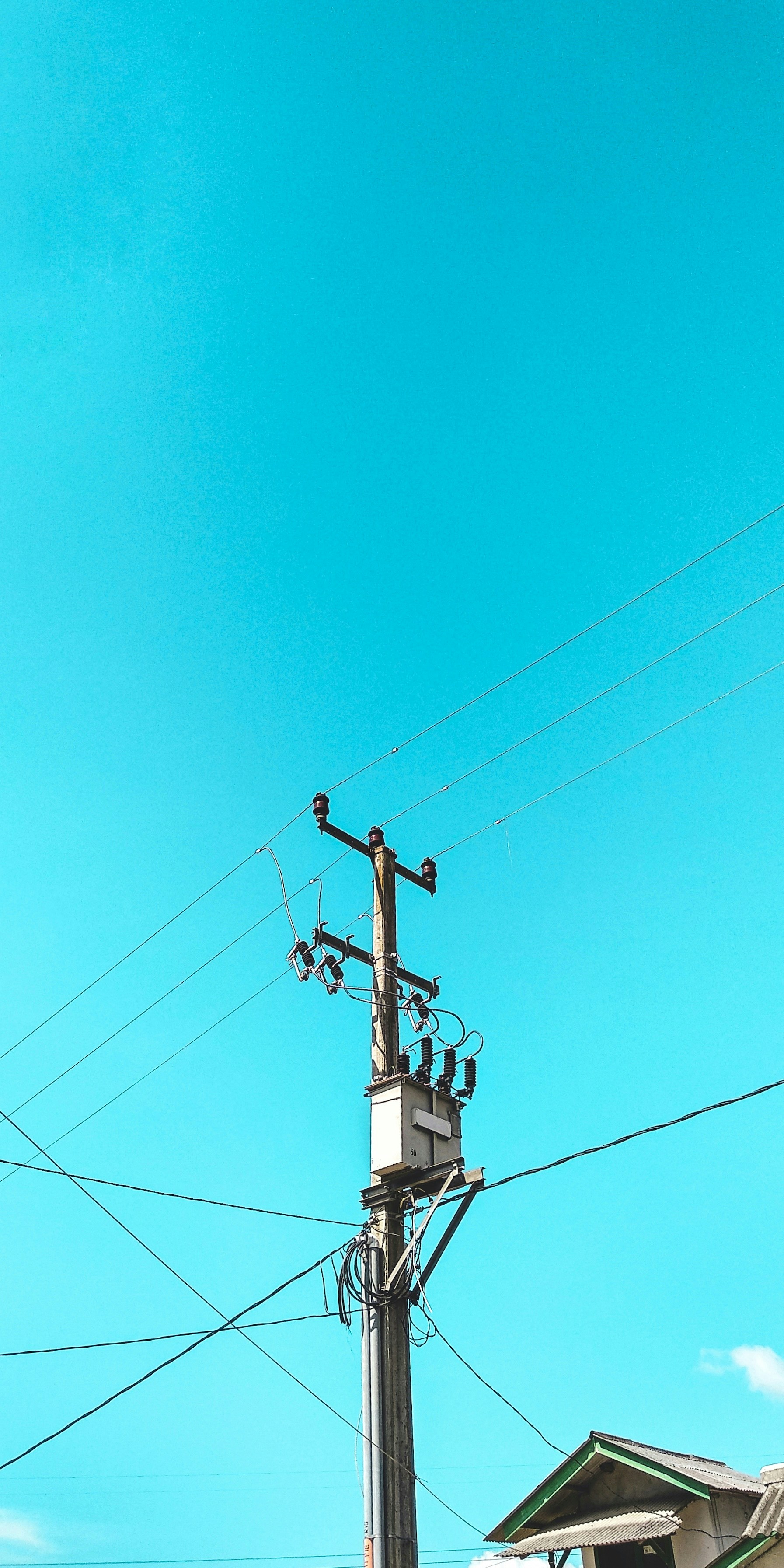 Electric utility pole with power lines stretching across a bright turquoise sky, juxtaposed with a nearby building. 