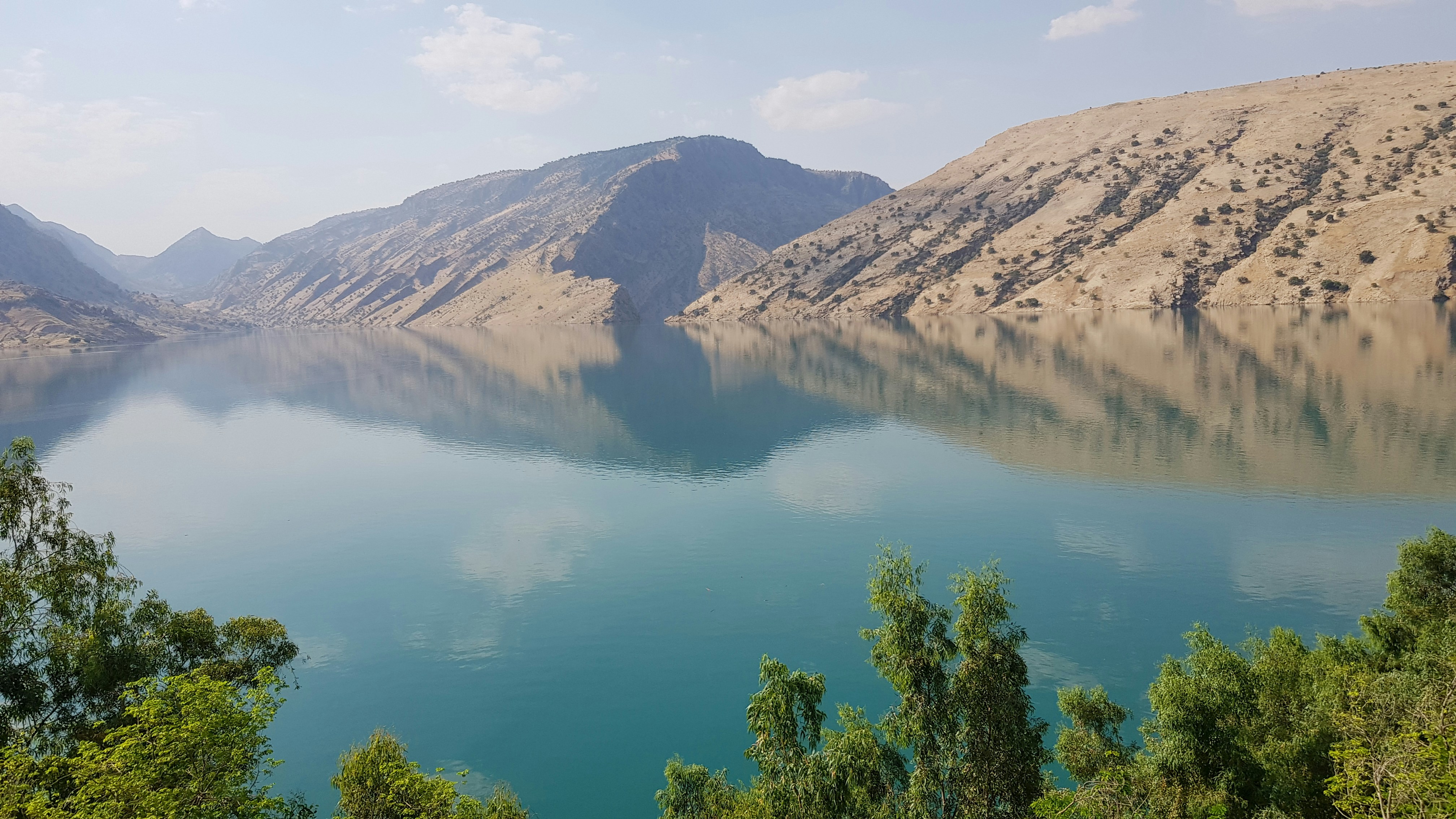 Tranquil lake reflecting surrounding mountains and sky, framed by lush greenery on the left. The scene captures the harmony of nature's elements.