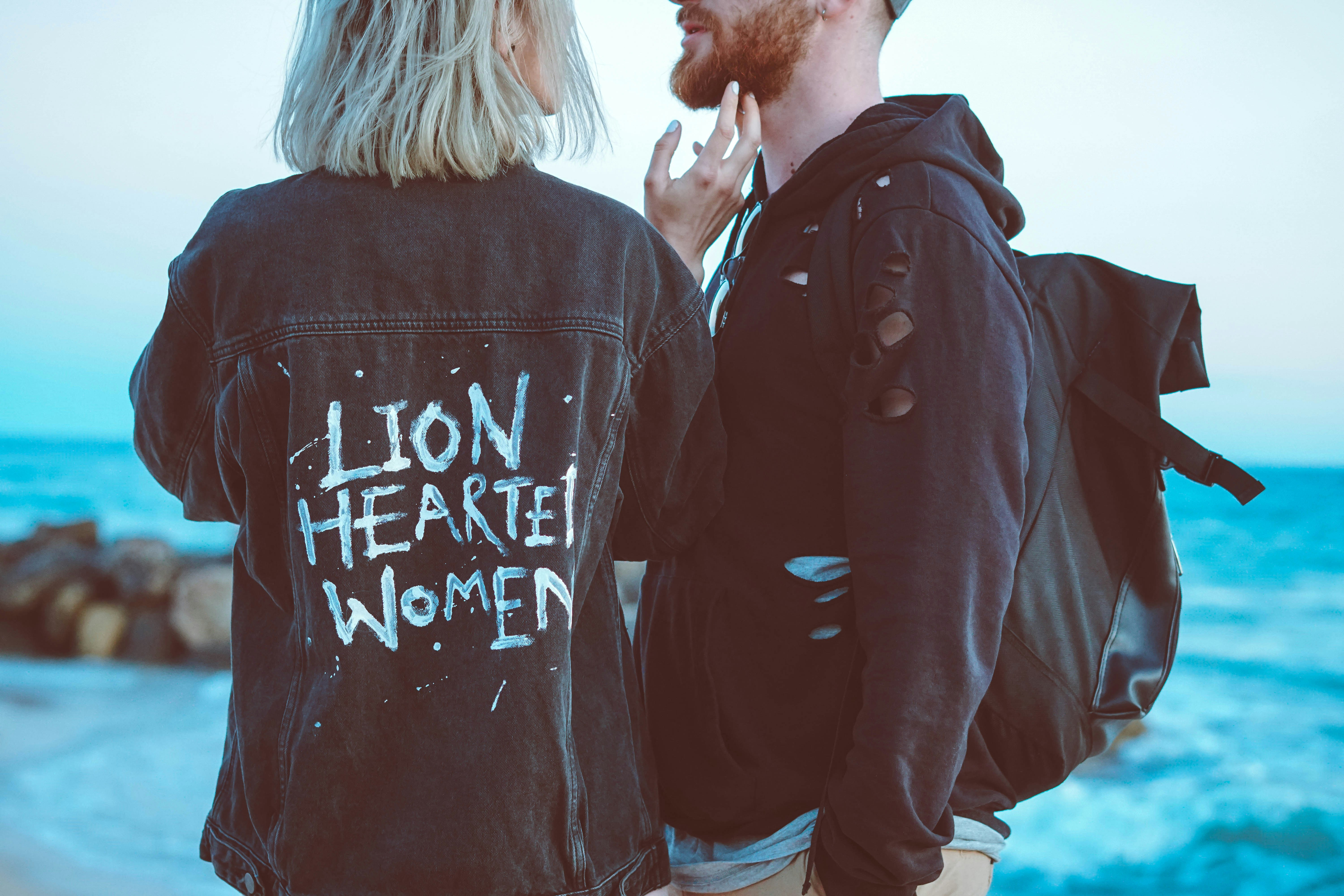 Couple sharing an intimate moment on the beach, with the woman's denim jacket displaying the phrase 'LION HEARTED WOMEN.'