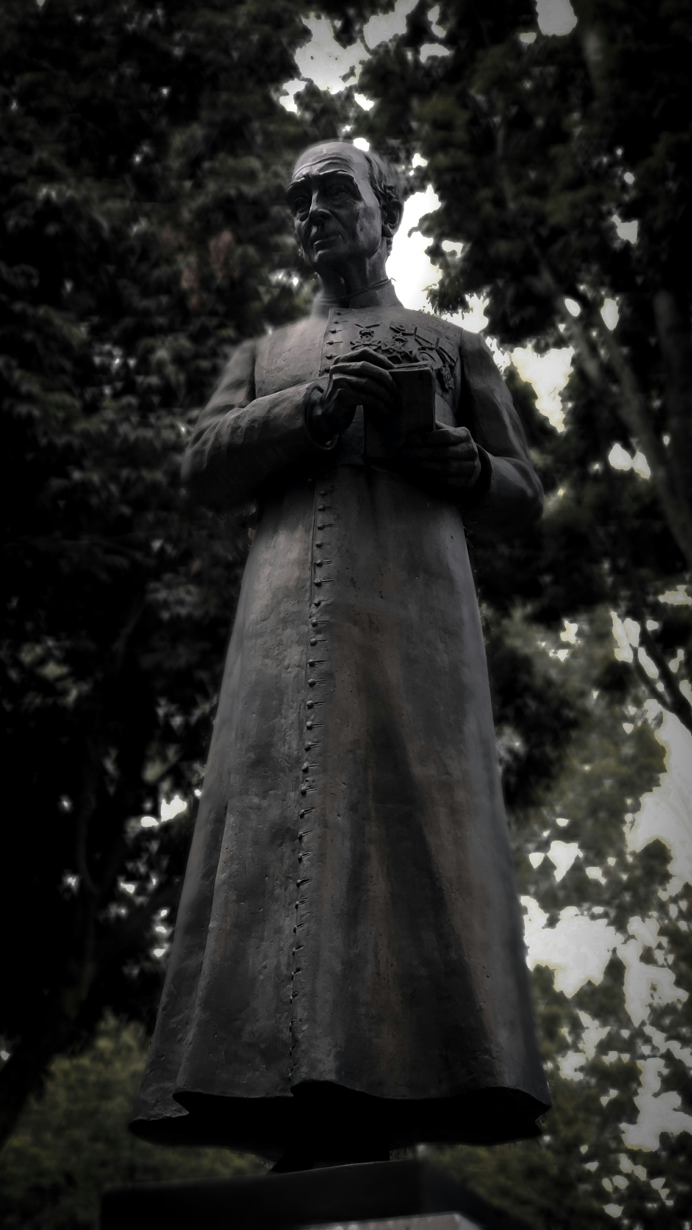 A bronze statue of a solemn figure holding a book, surrounded by lush greenery. The statue exudes an air of wisdom and reflection.