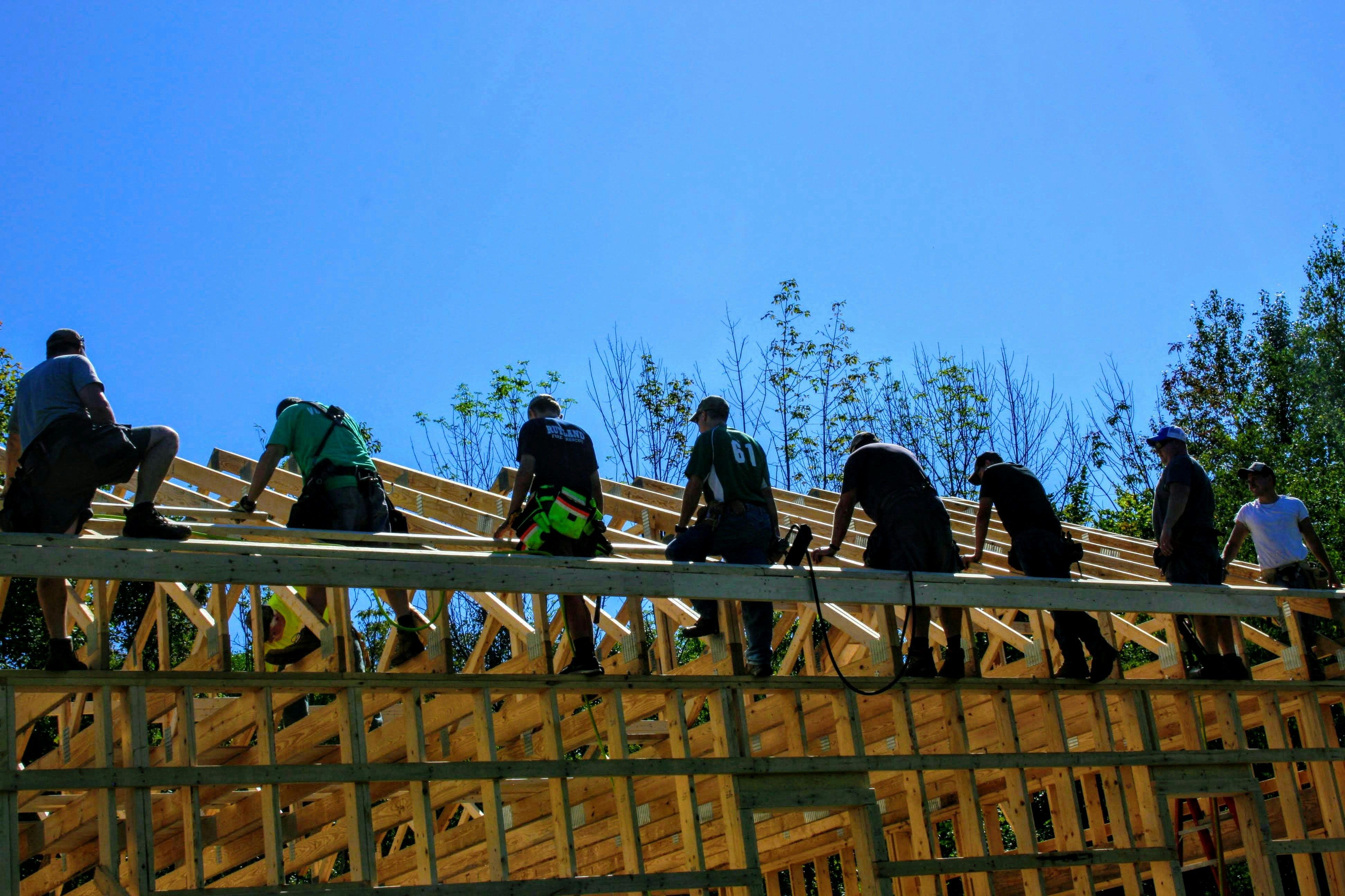 Construction workers collaborating on a wooden roof framework under a clear blue sky.