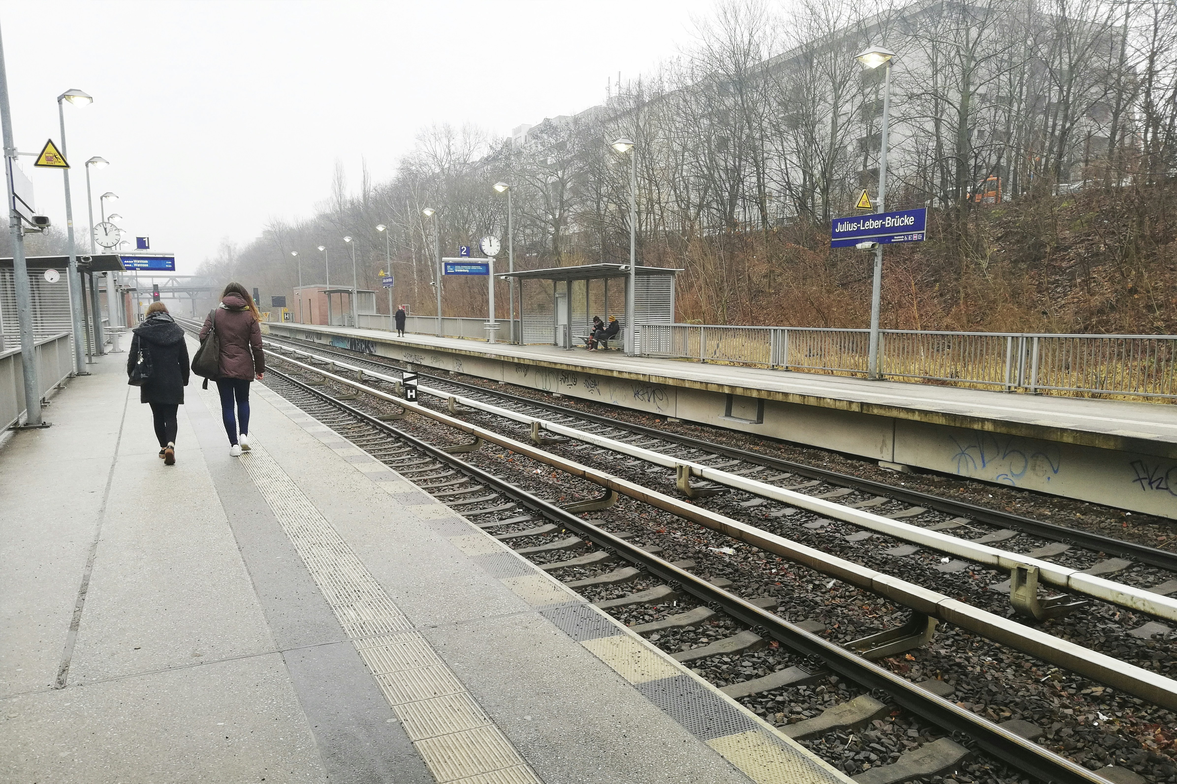 Two pedestrians walking along a nearly empty train platform on a foggy day, with distant figures seated and train tracks visible.