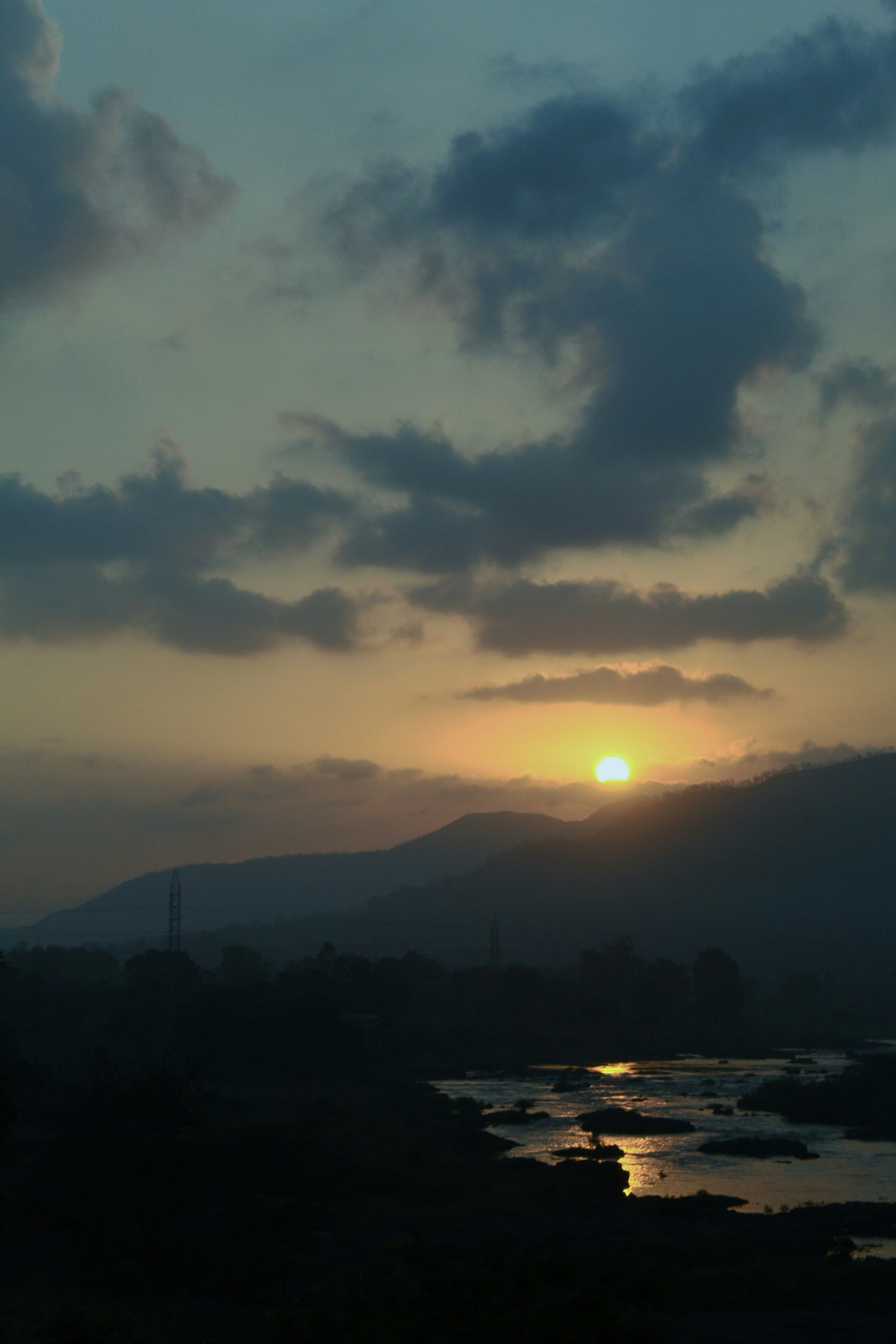 Golden sun setting behind distant hills, casting reflections on a winding river below. The sky is adorned with soft clouds, creating a serene atmosphere.