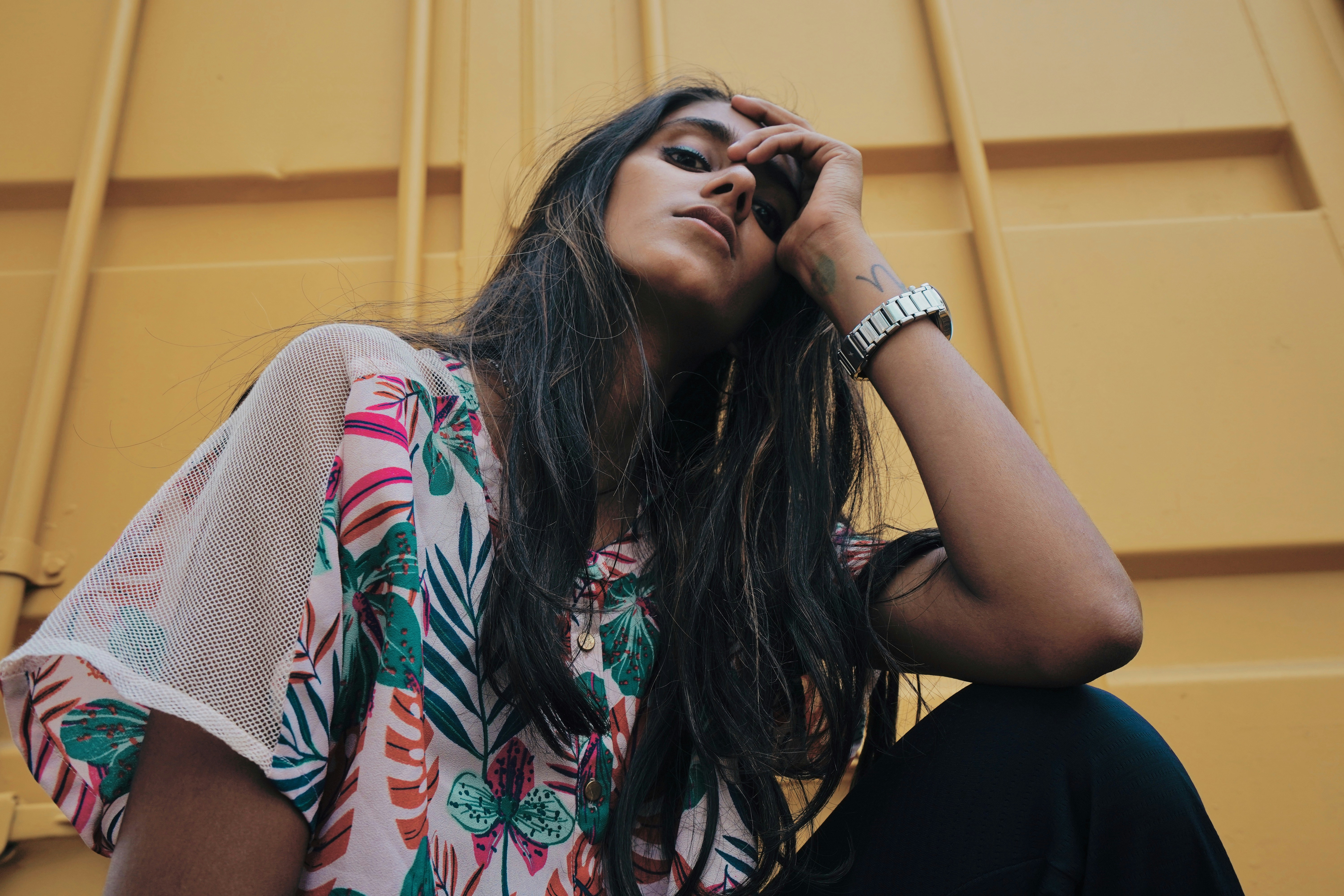 Woman in a floral shirt poses thoughtfully against a yellow geometric wall.