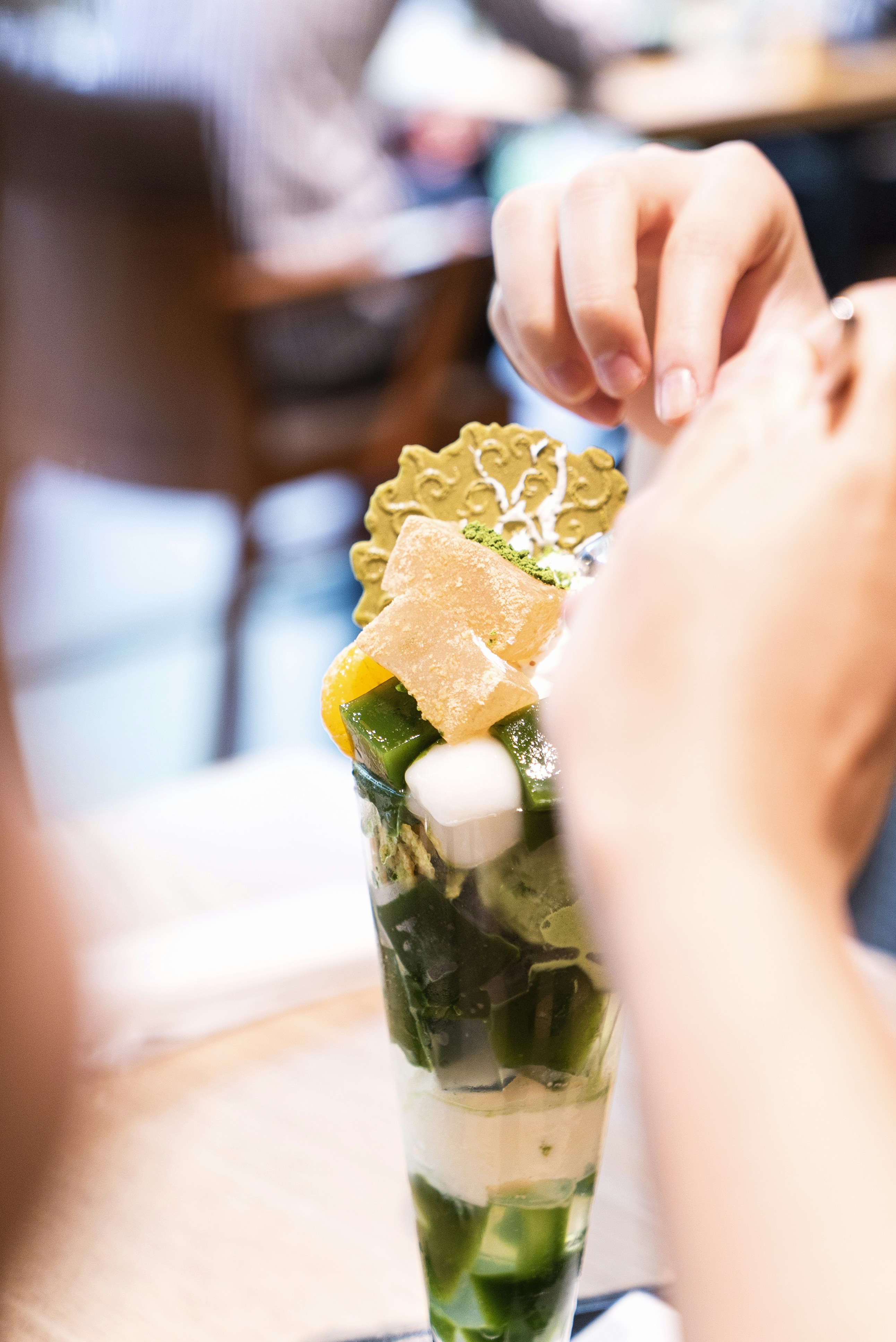 Layered dessert featuring green jelly, mochi, and decorative elements, elegantly presented in a tall glass. The scene captures a moment of enjoyment as a hand reaches for a treat.