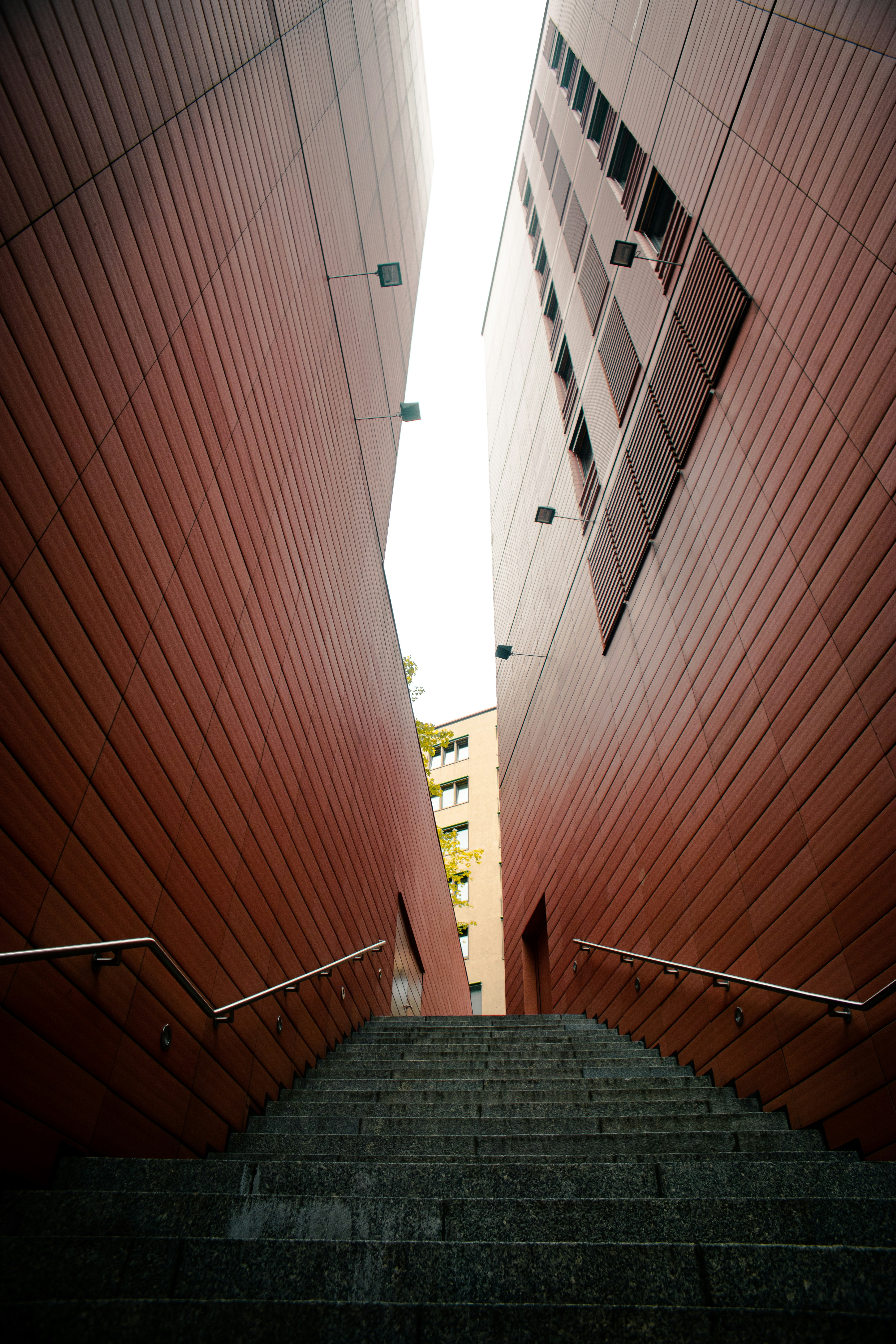 Staircase flanked by tall, red wooden walls leading upward towards a bright sky. The geometric lines create a striking perspective.