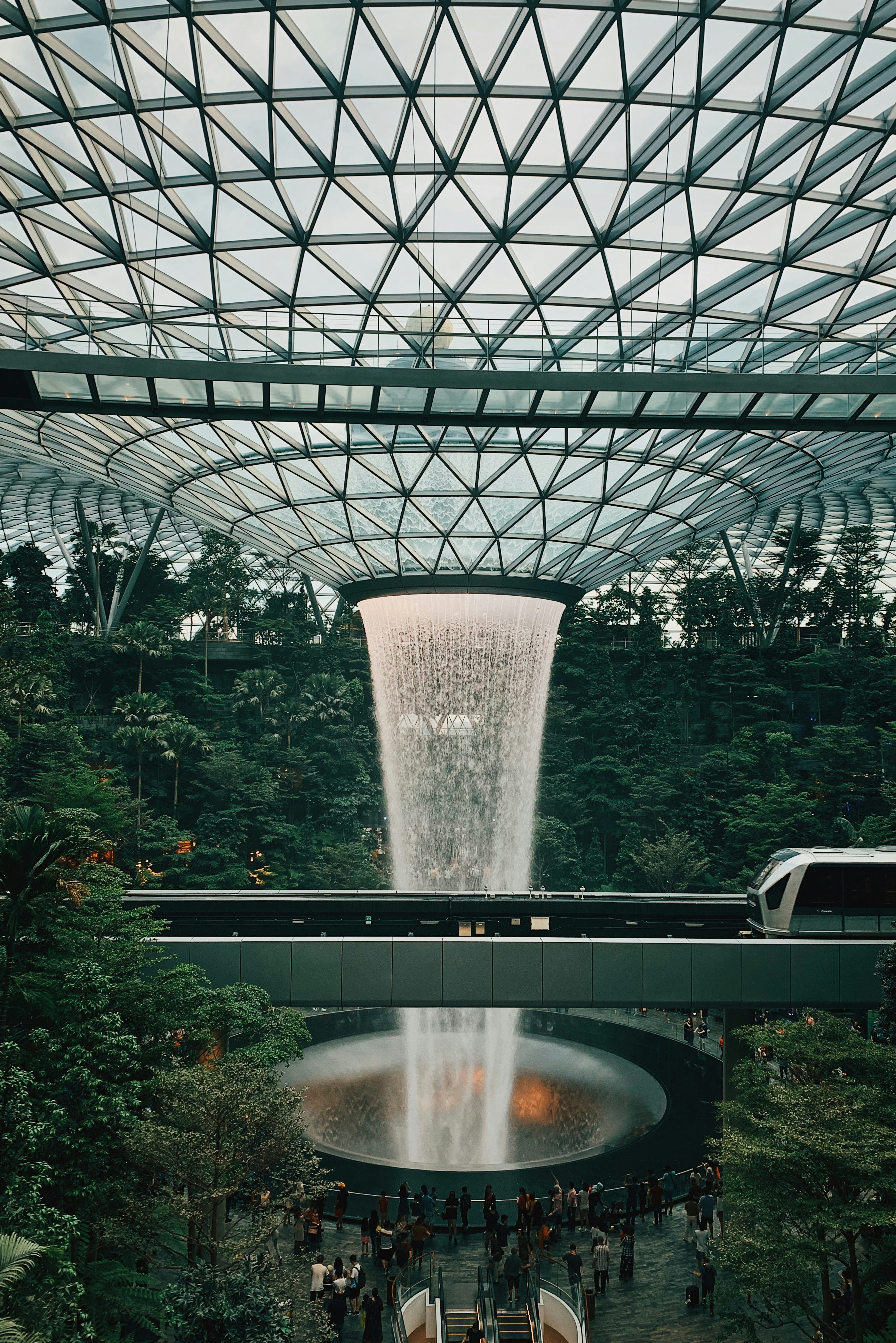 A breathtaking indoor waterfall cascades from a geometric glass ceiling, surrounded by lush greenery and curious visitors. The scene captures the fusion of natural and built environments.