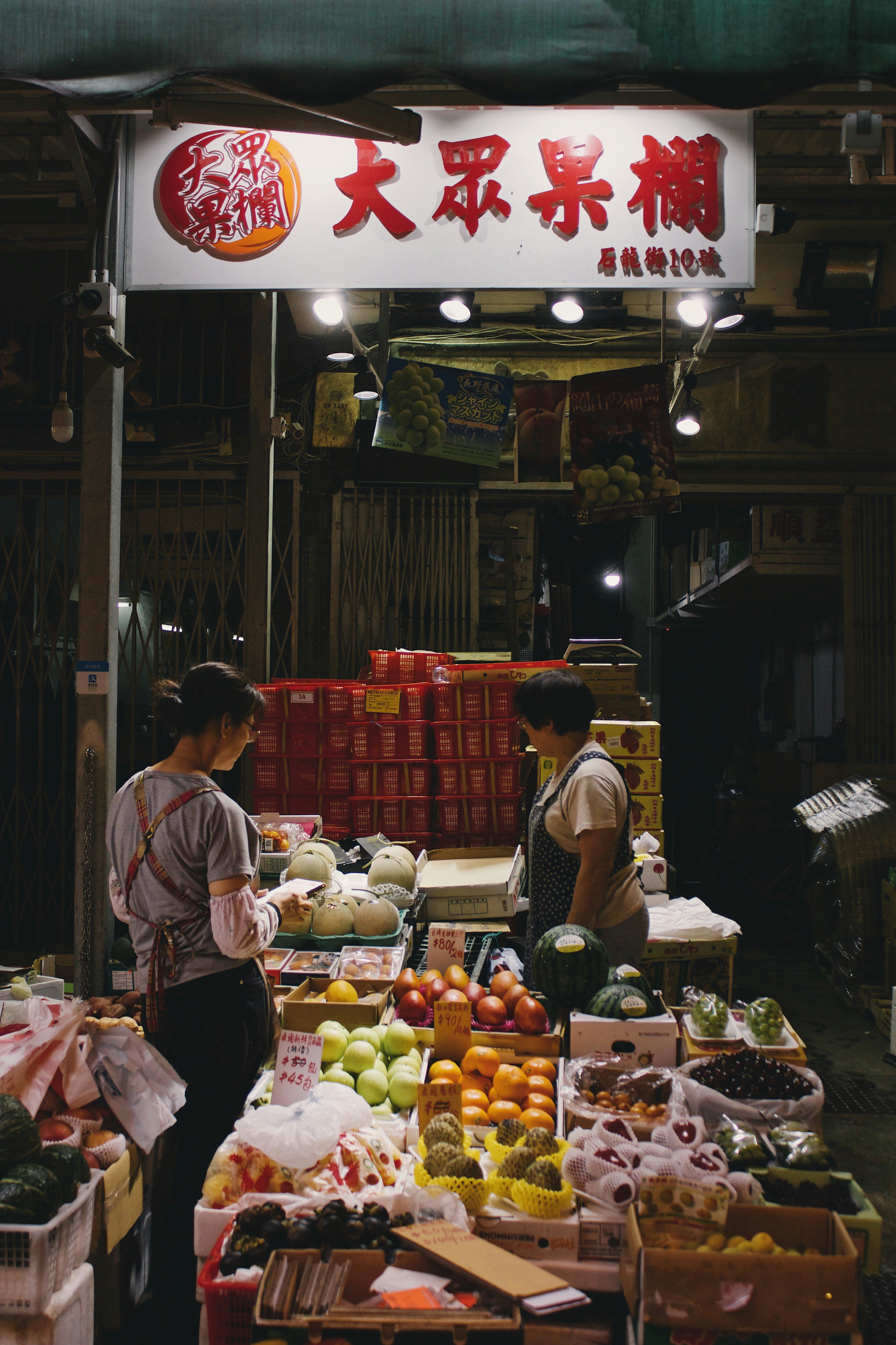 Vibrant night market scene featuring two individuals selecting fresh produce amidst a colorful display of fruits and vegetables. A glowing sign adds to the ambiance.