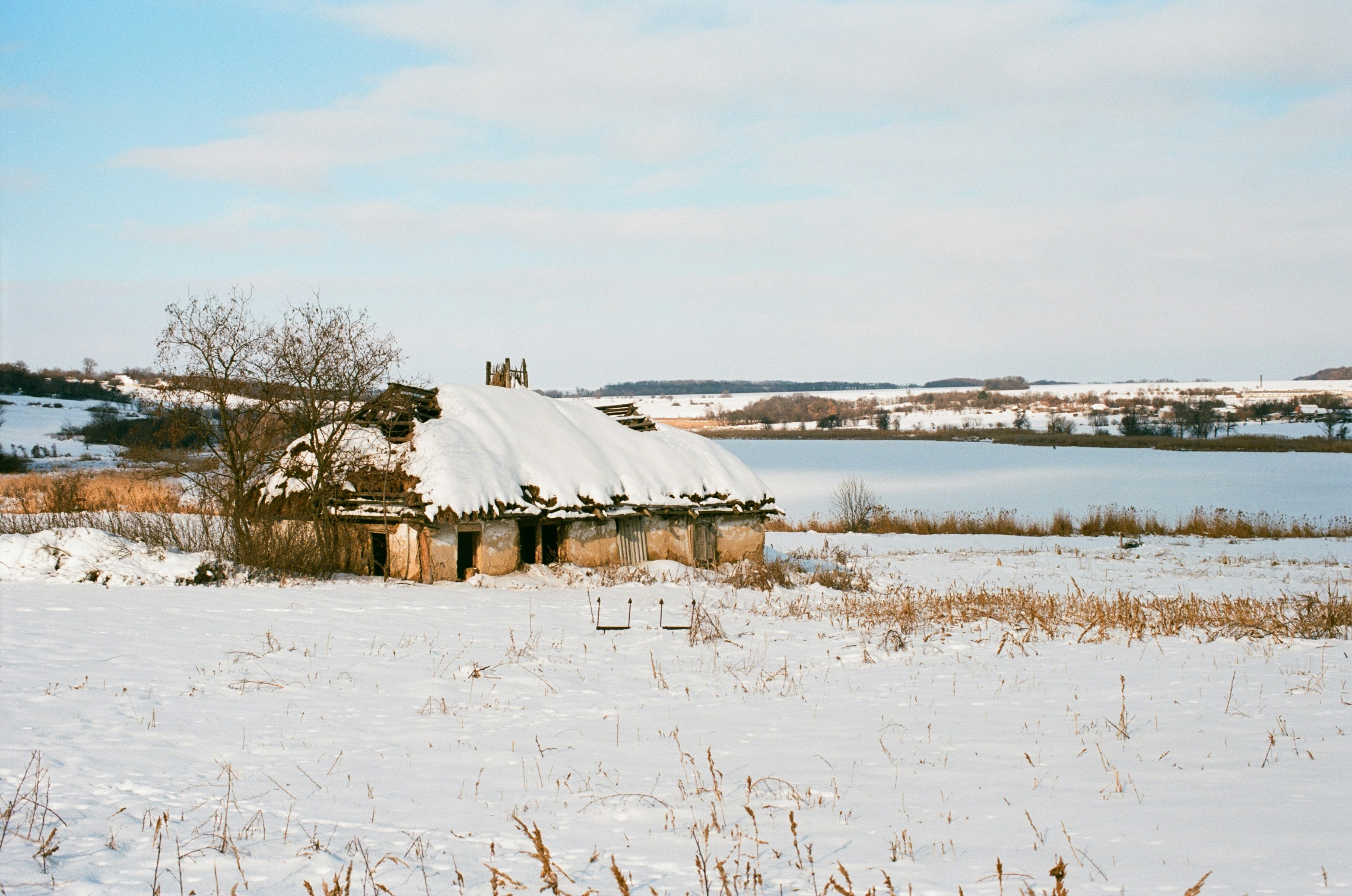 Abandoned cottage with a snow-covered roof beside a tranquil lake, surrounded by frosty grasslands and distant hills.