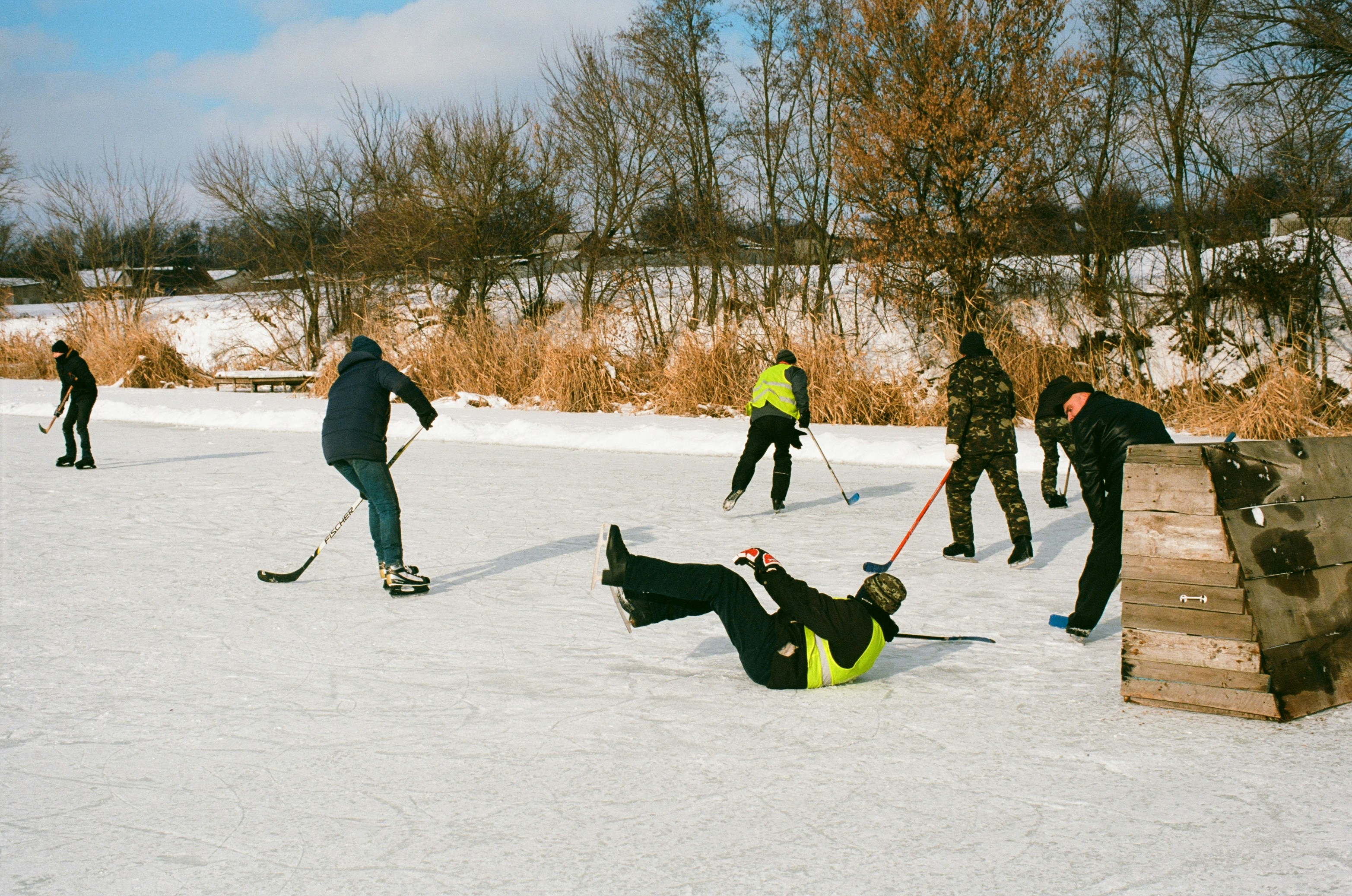 A winter ice-skating scene captured in a photograph on a frozen pond with several skaters, including a figure lying mid-fall near the center. The scene shows a wooden ramp and bare trees framing the action.
