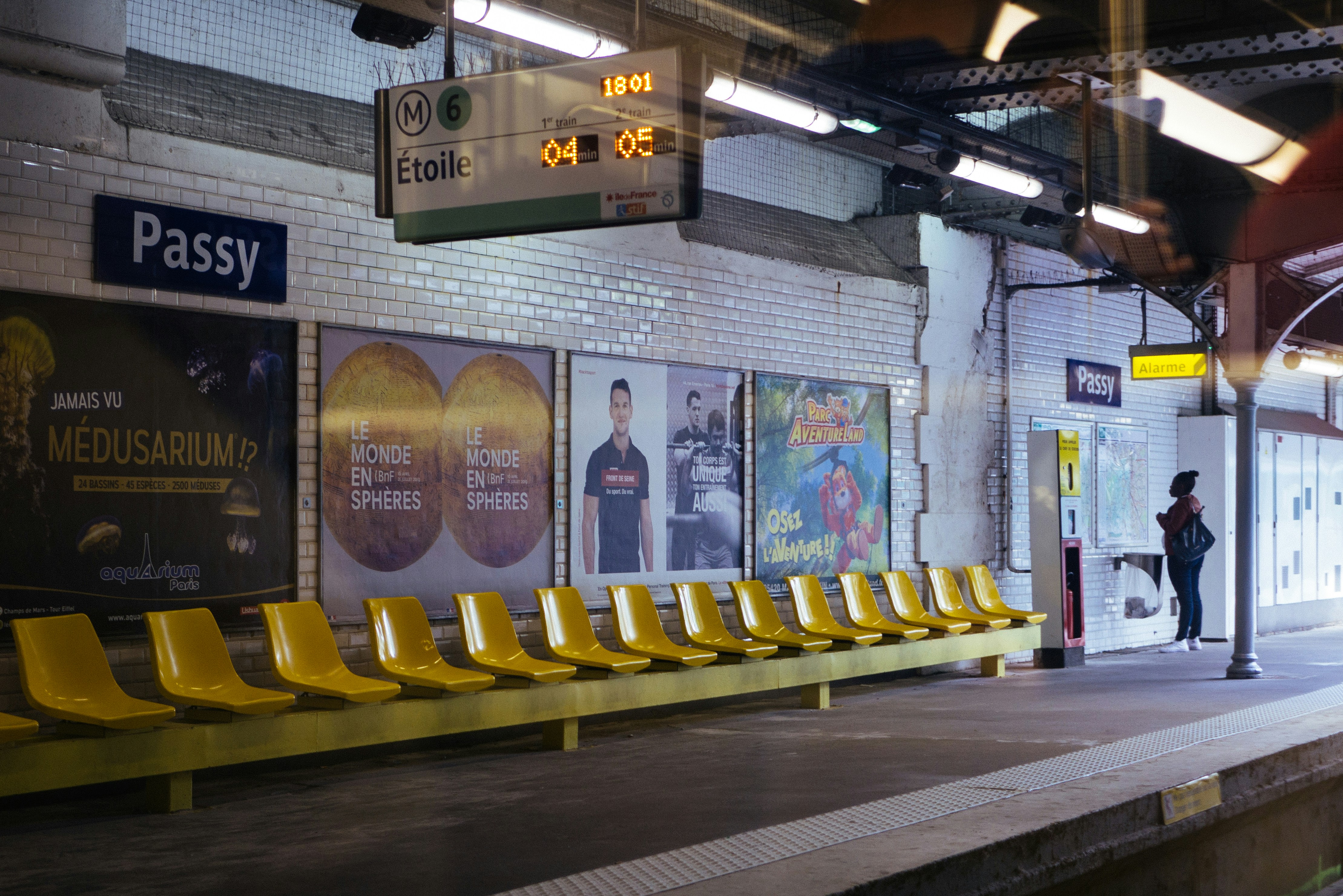 A quiet subway platform featuring bright yellow seats, with advertisements and a lone figure waiting for the train. The ambiance reflects a moment of stillness in a bustling city.