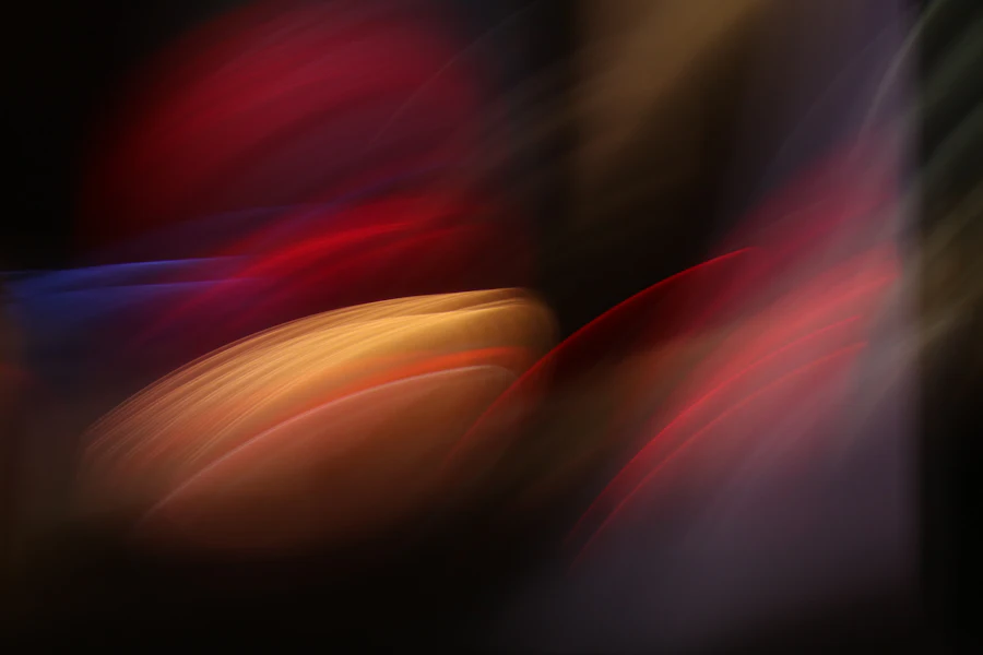 Abstract blur of red, gold, blue, and purple light streaks against a dark background. The warm oval shape near the lower left draws the eye amid sweeping motion trails.