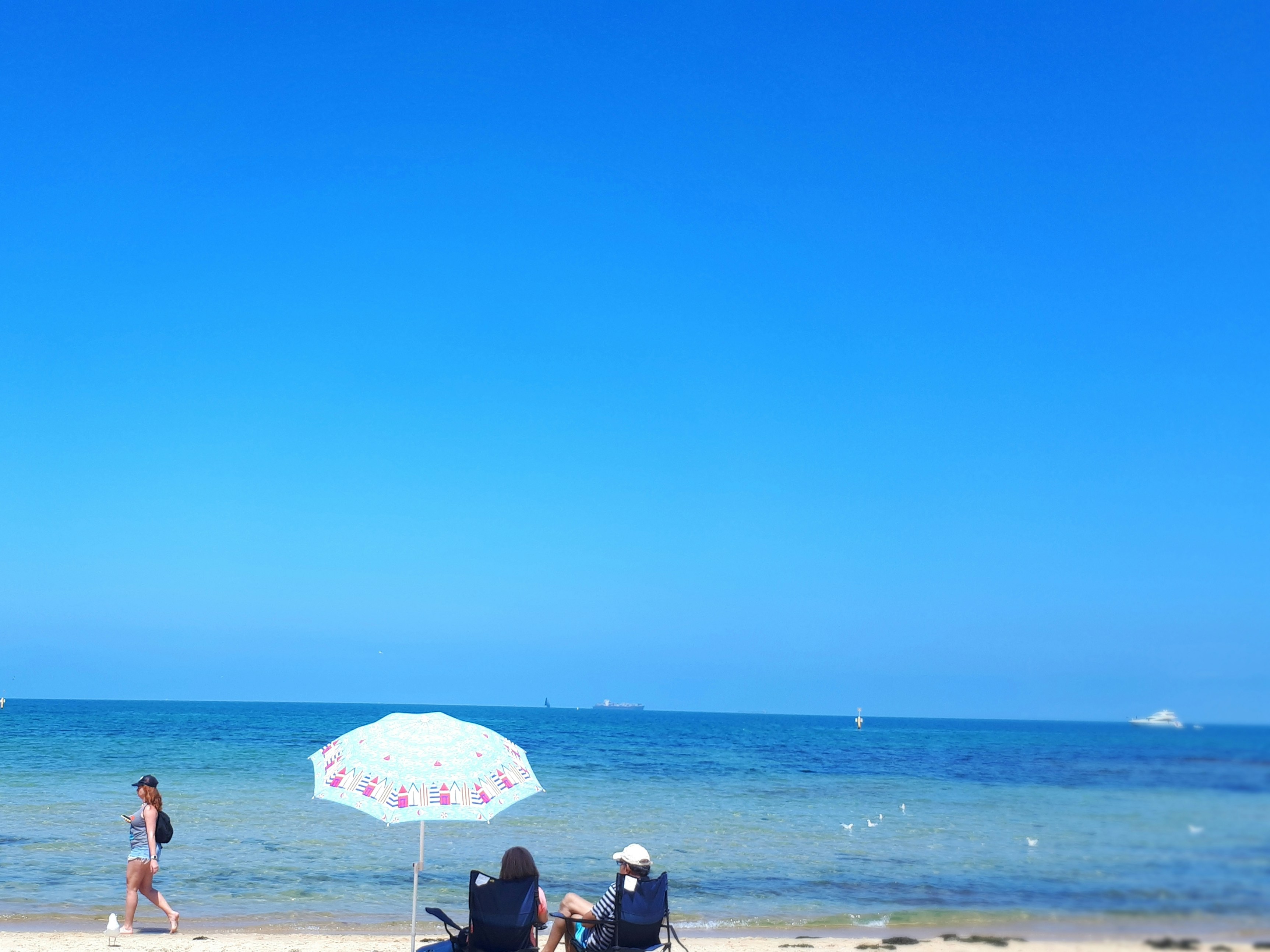 Beachgoers relax under a colorful umbrella while a passerby strolls along the shore, capturing a tranquil summer day.