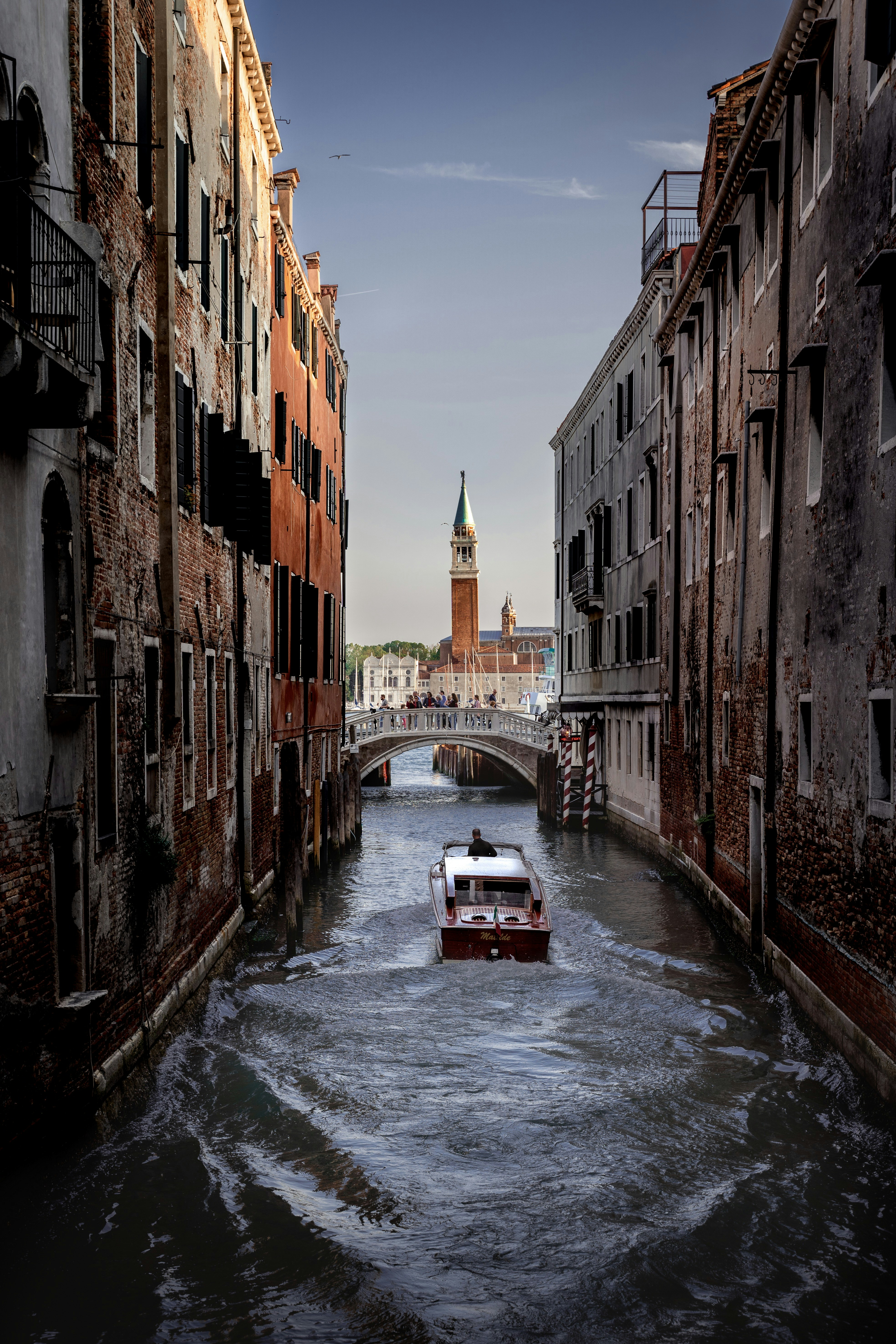A motorboat glides through a narrow Venetian canal, flanked by historic buildings, with the iconic campanile in the background.