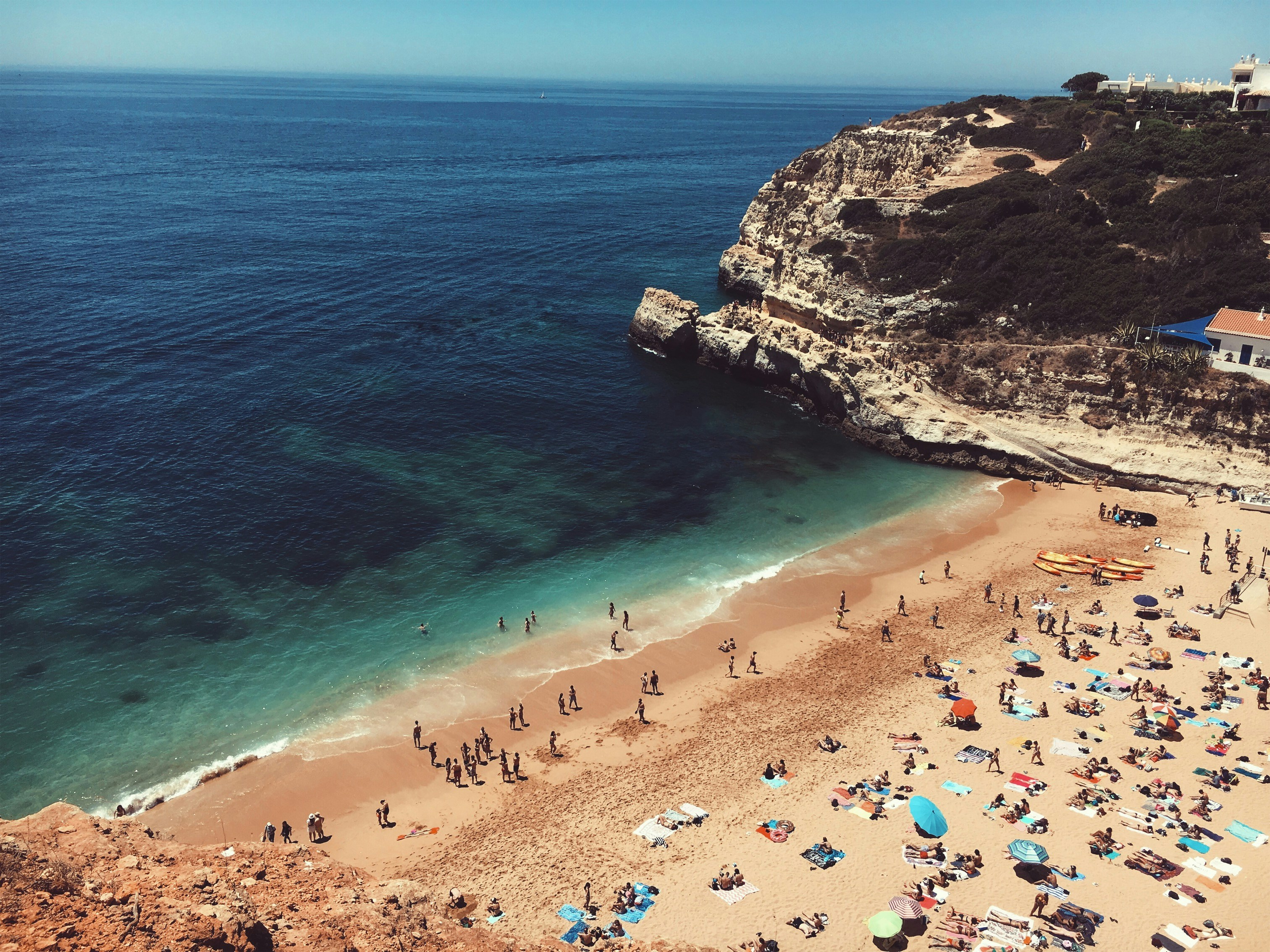 Crowded sandy beach bordered by turquoise waters and rugged cliffs under a clear sky.
