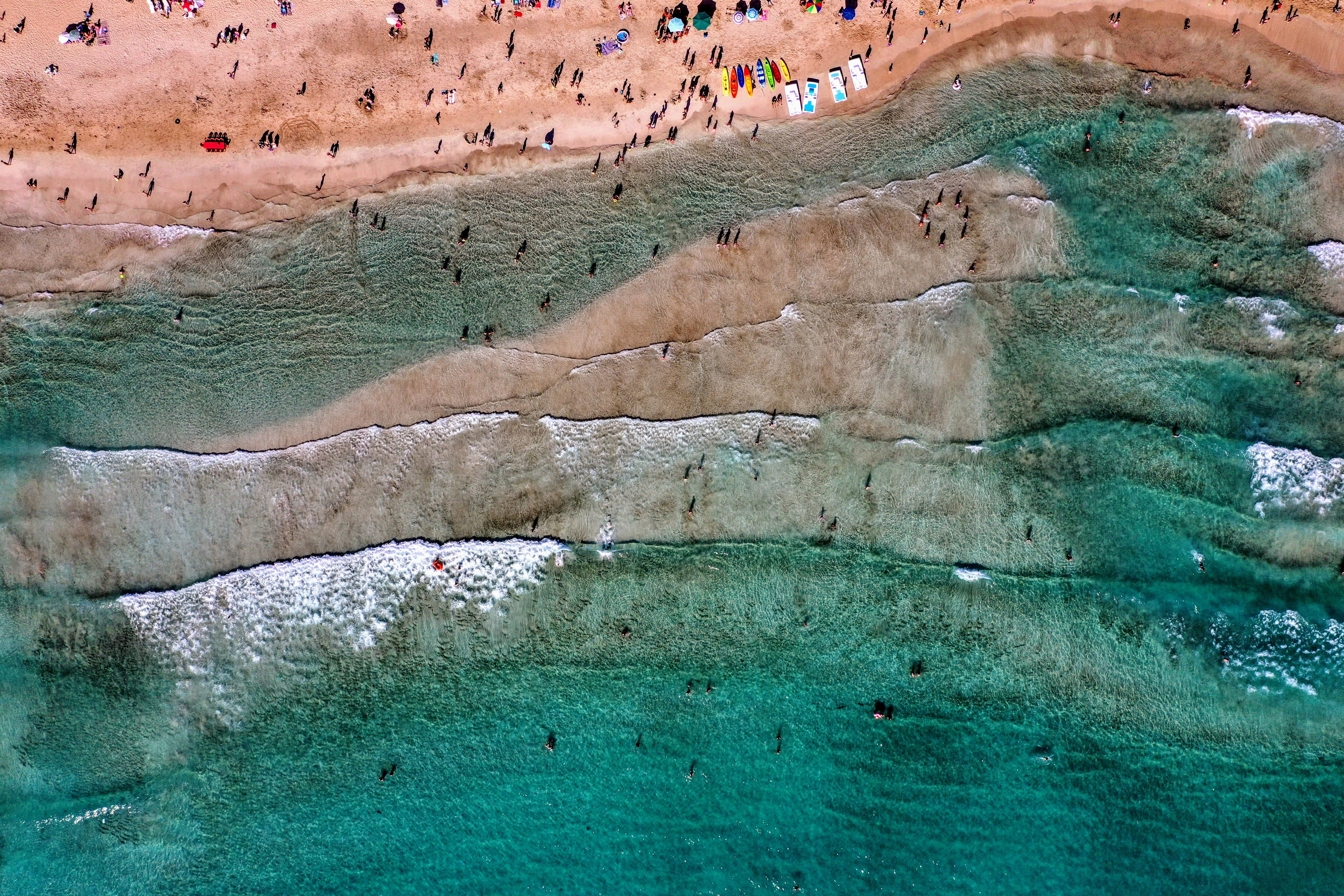 Aerial view of turquoise waves gently lapping against a sandy shore with scattered beachgoers.