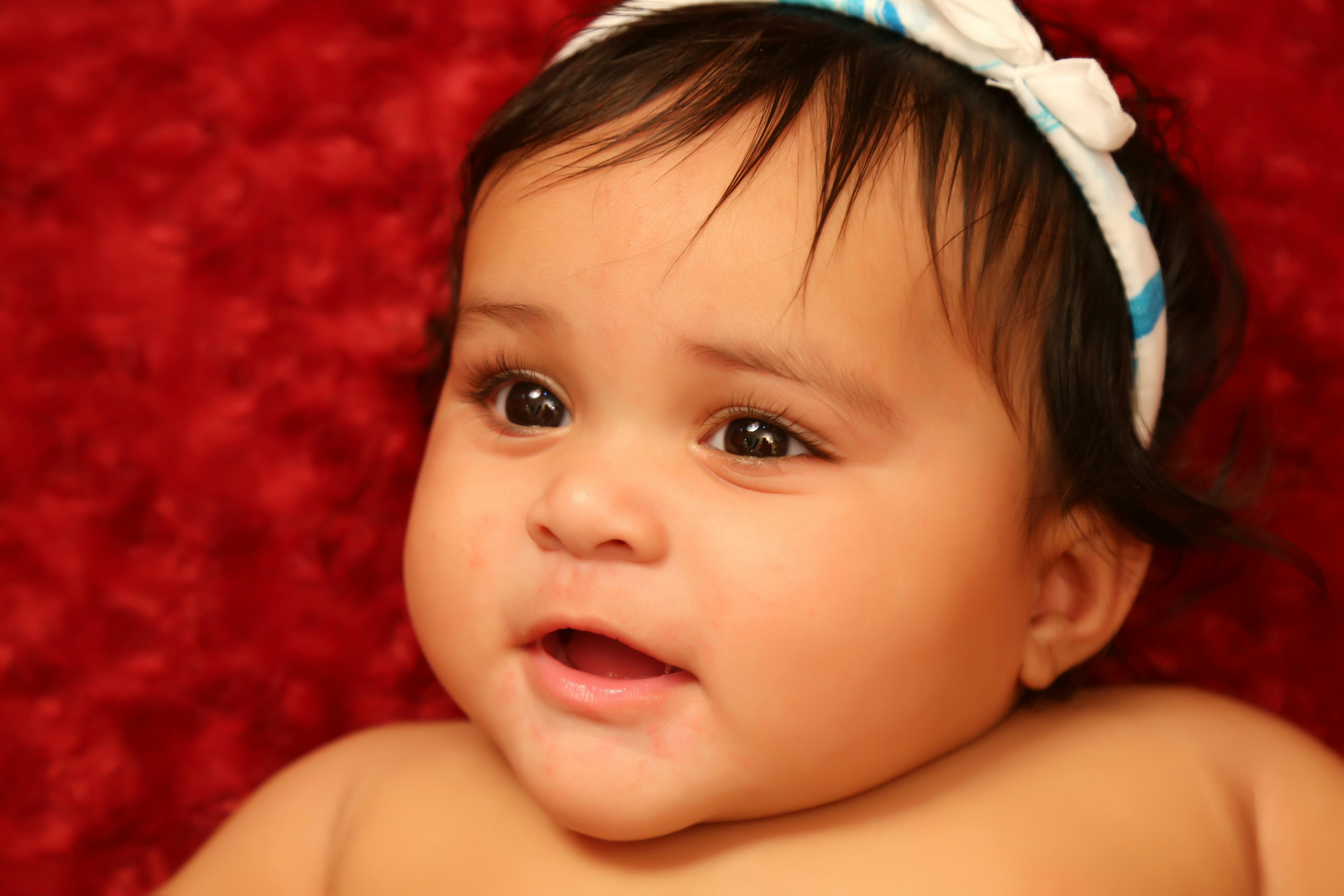 Infant with a playful expression and a stylish headband, resting against a plush red backdrop.