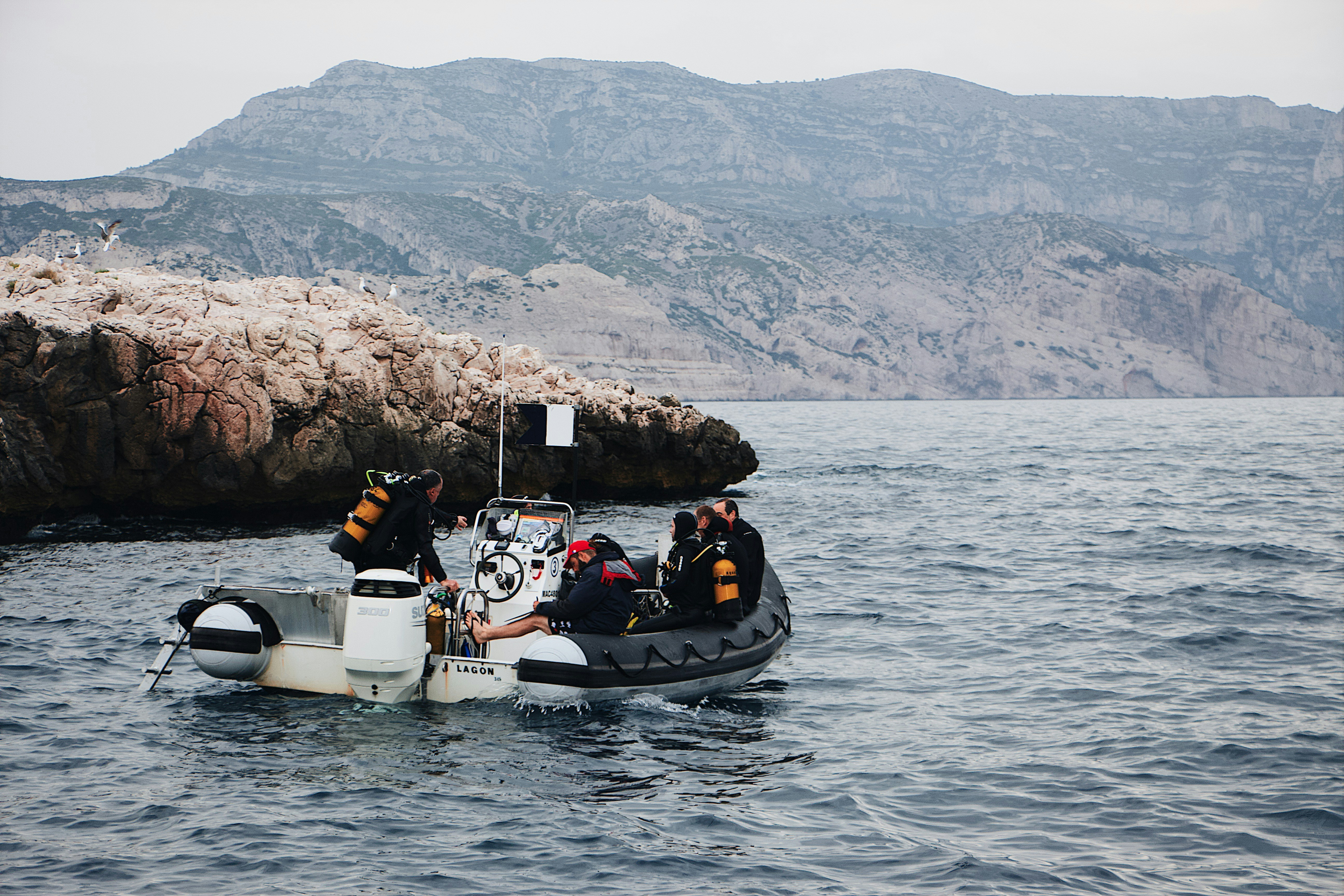 Group of individuals aboard a boat near rocky coastline, preparing for exploration on the water. The backdrop features rugged mountains under a cloudy sky.