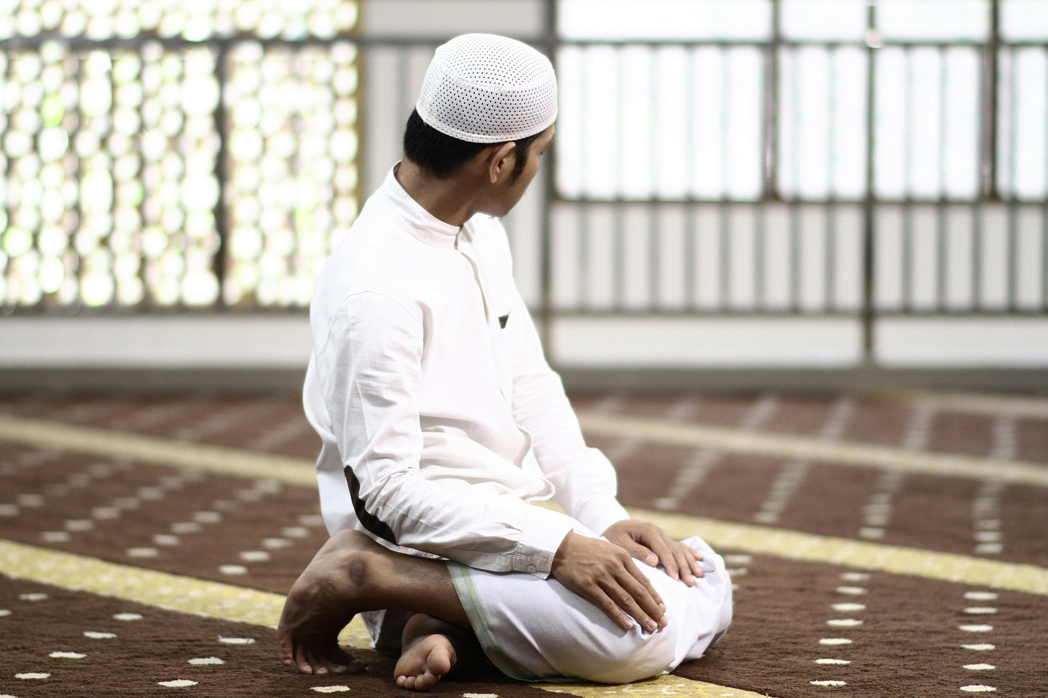A man in traditional attire sits in a mosque, gazing thoughtfully away from the camera, surrounded by intricate patterns on the floor.