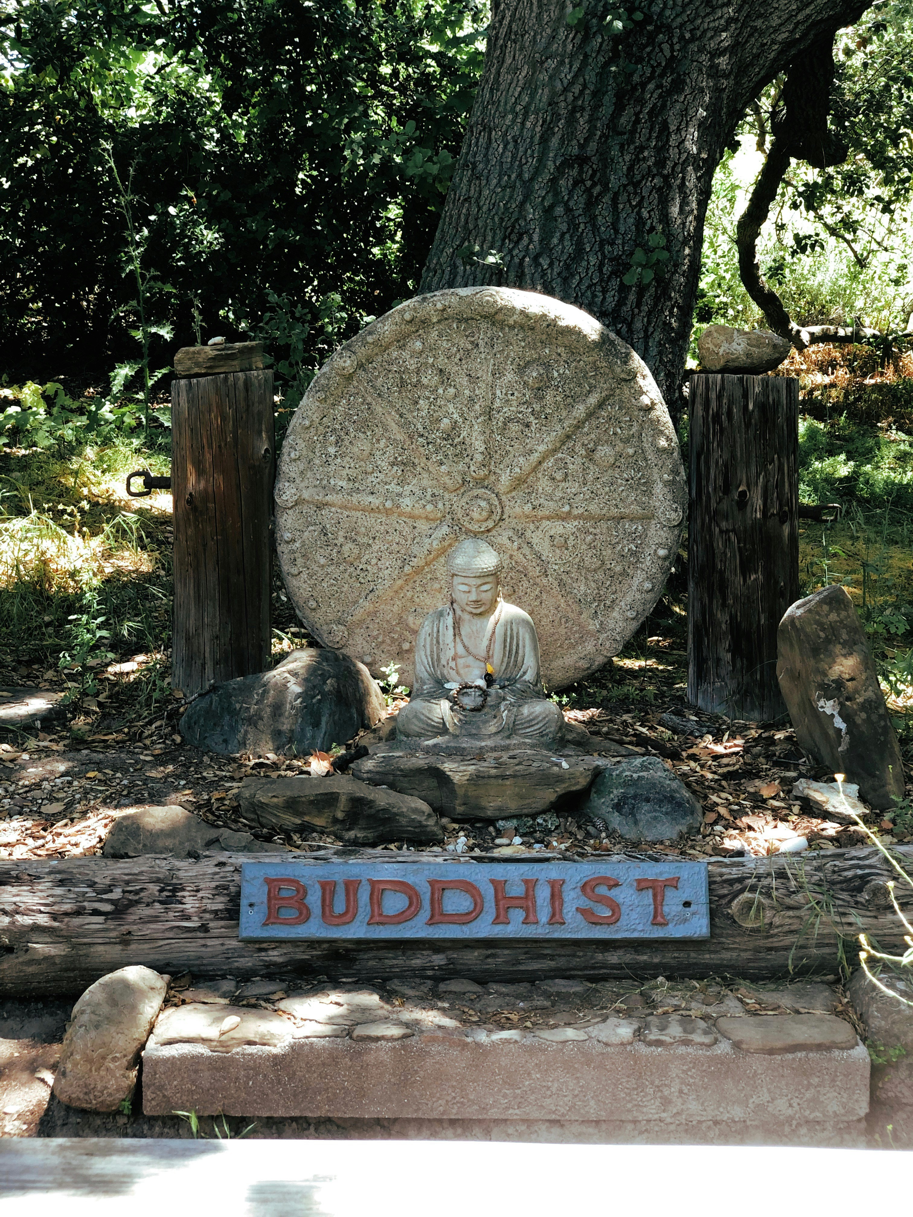 A serene Buddhist statue sits beneath a large stone wheel, surrounded by natural elements in a tranquil setting.