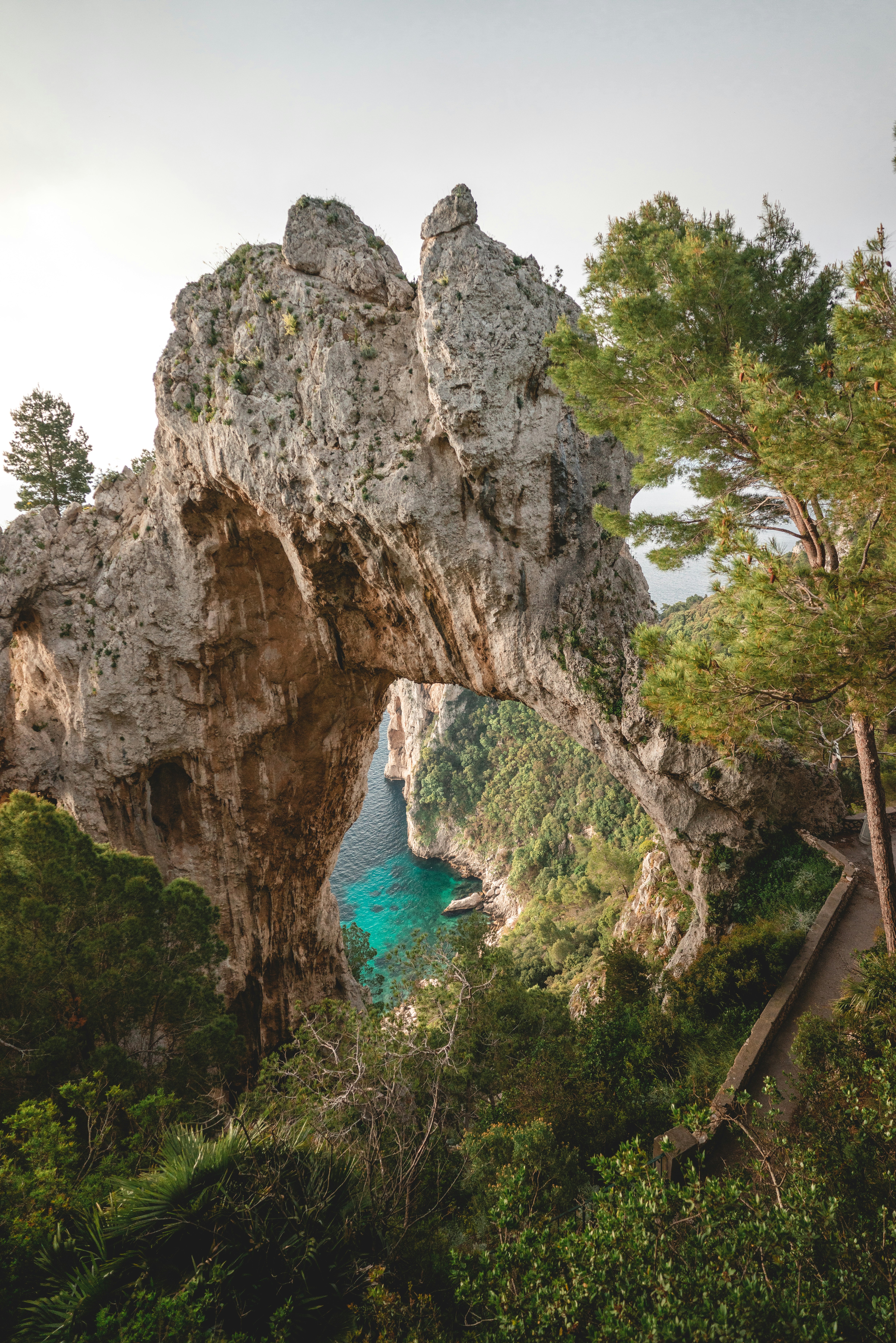 Majestic rock arch framing a serene turquoise sea, surrounded by lush greenery and rugged cliffs.