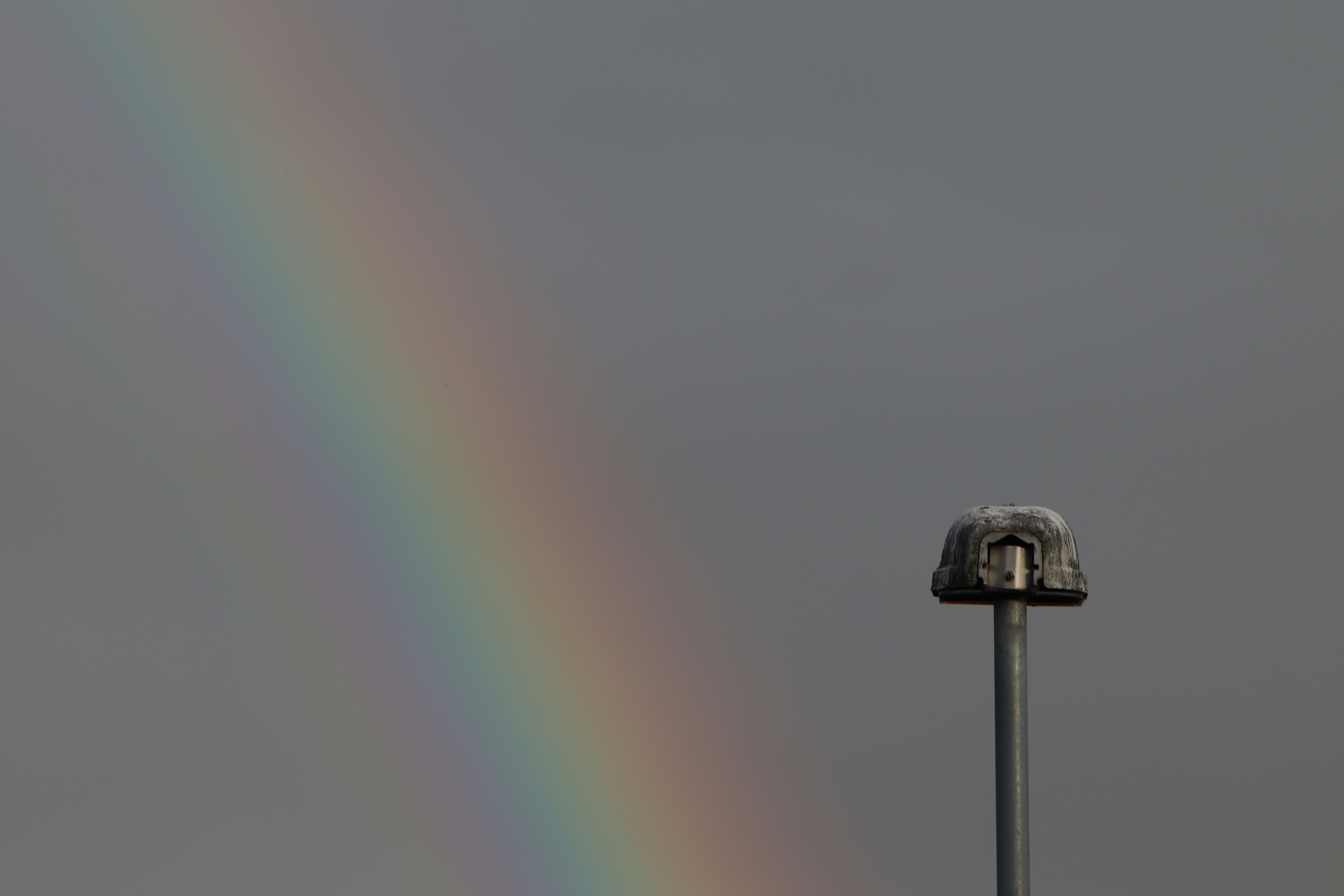 A weathered light fixture stands tall against a backdrop of a vibrant rainbow emerging from dark clouds.