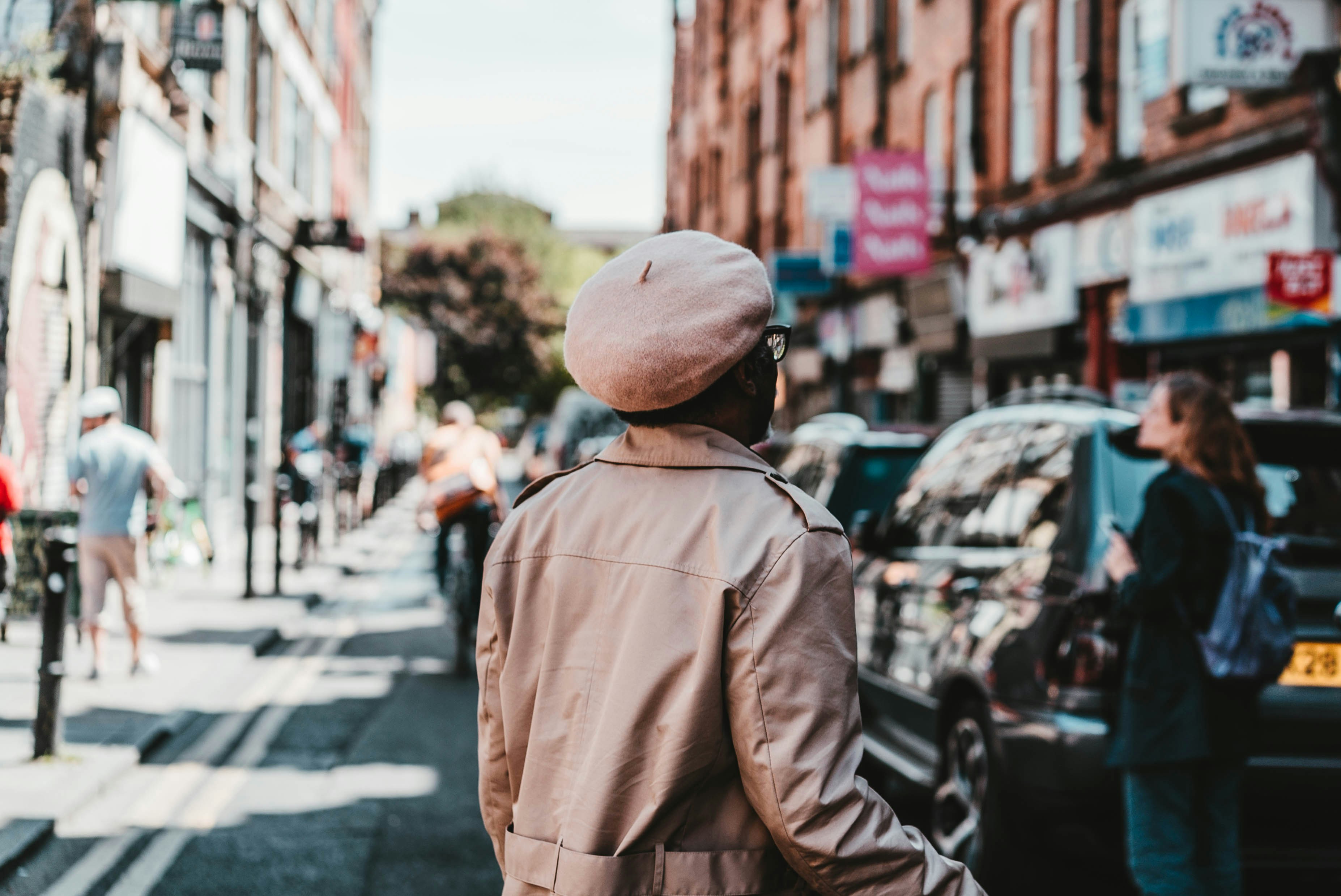 Person in a trench coat and beret walks down a bustling city street lined with shops and cars.