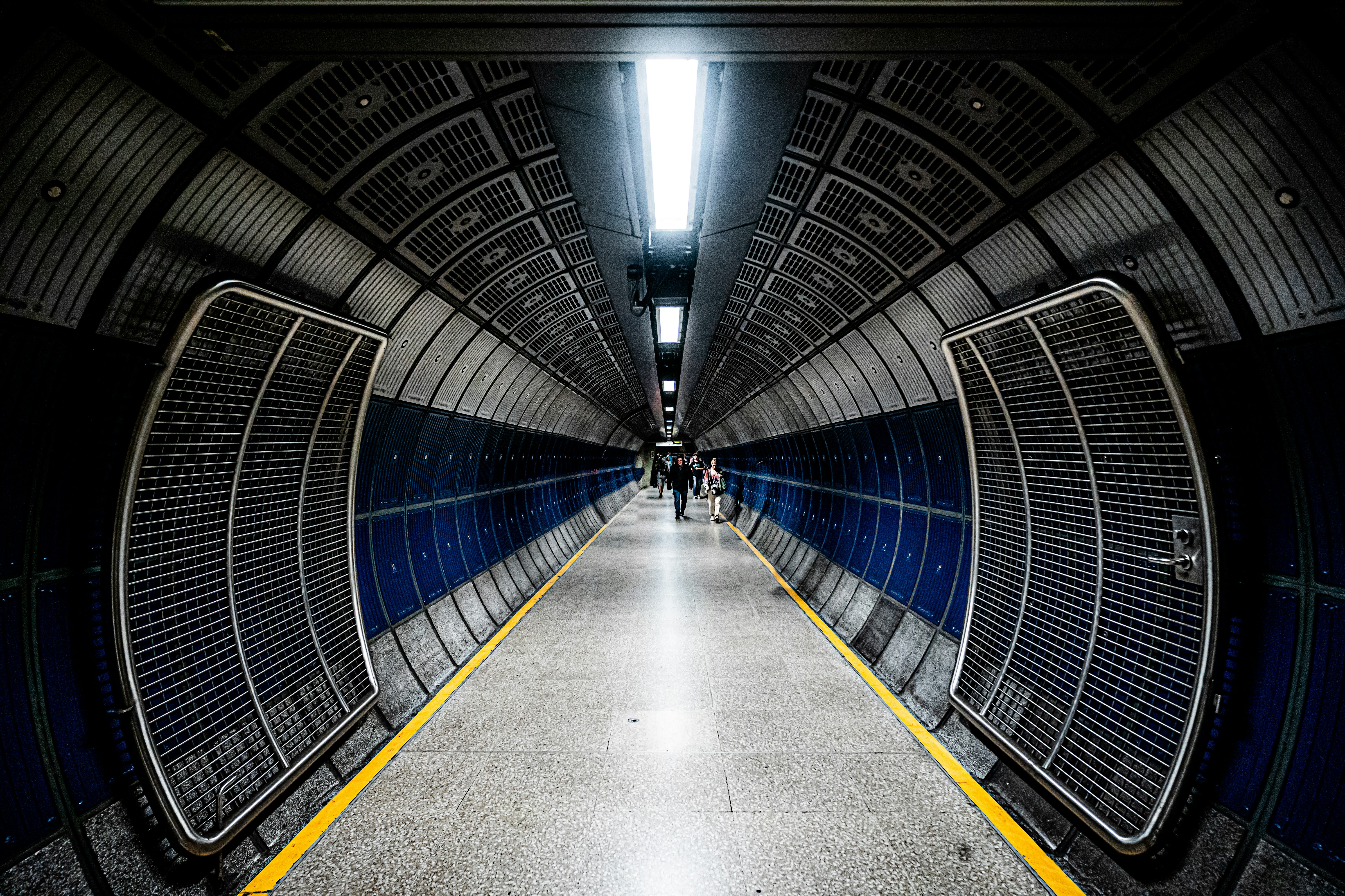 People walking through a futuristic, curved tunnel with blue panels and overhead lighting.