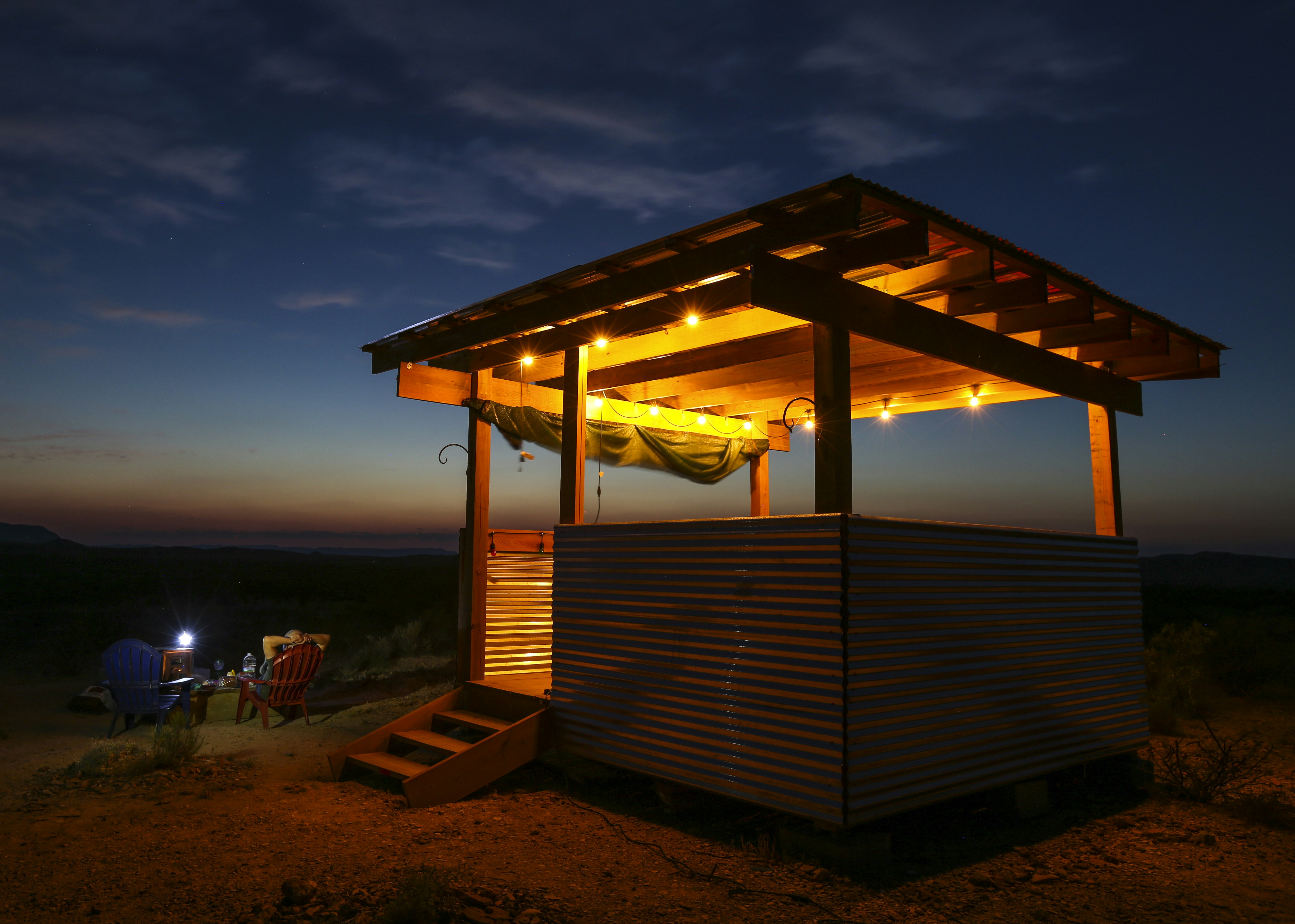 A cozy shelter illuminated by warm lights stands against the twilight sky, with a hint of outdoor seating nearby. The serene desert landscape surrounds the structure.