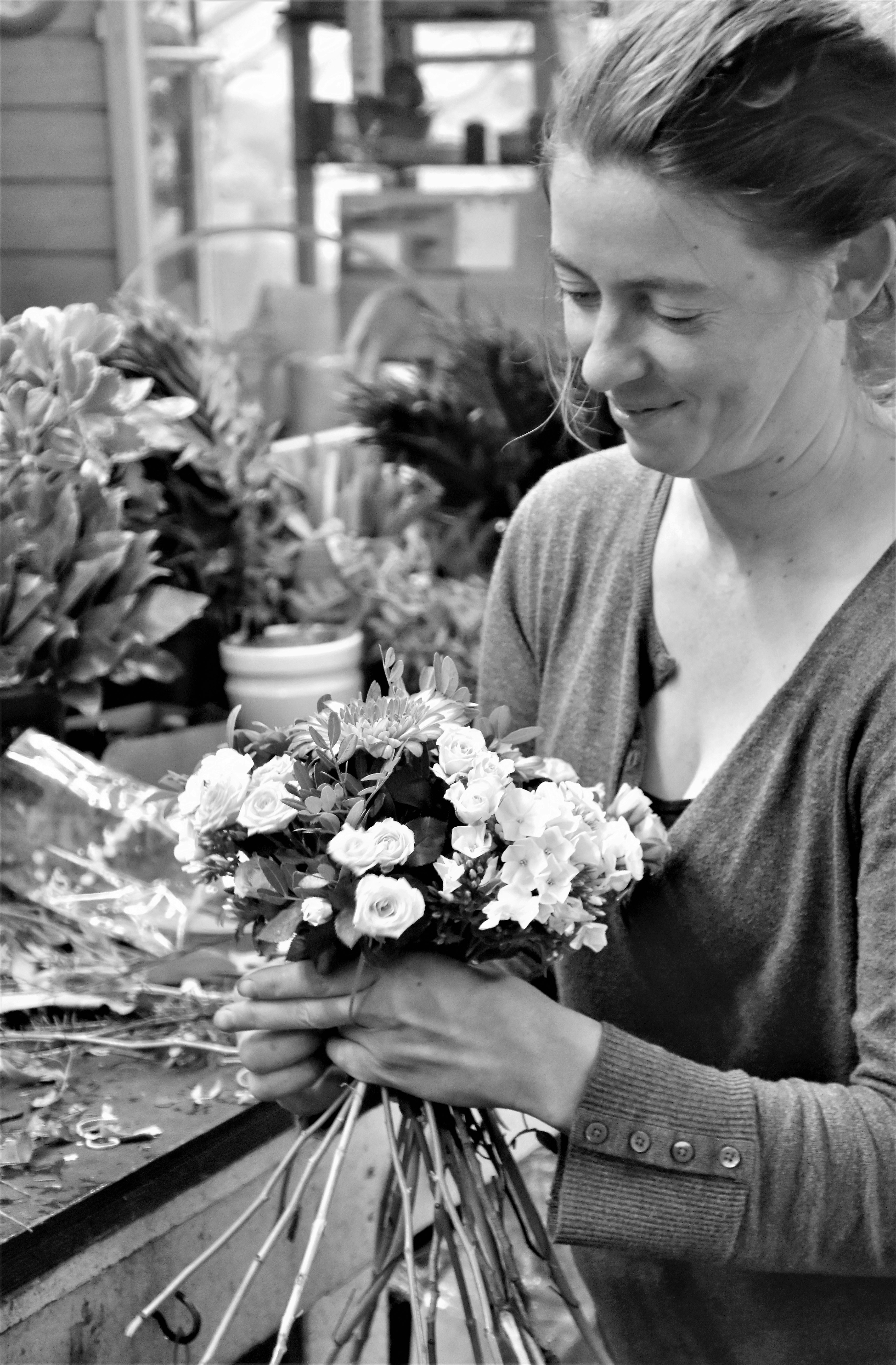 Florist arranging a bouquet of fresh flowers in a workshop filled with greenery and tools. The scene captures the artistry and dedication involved in floral design.