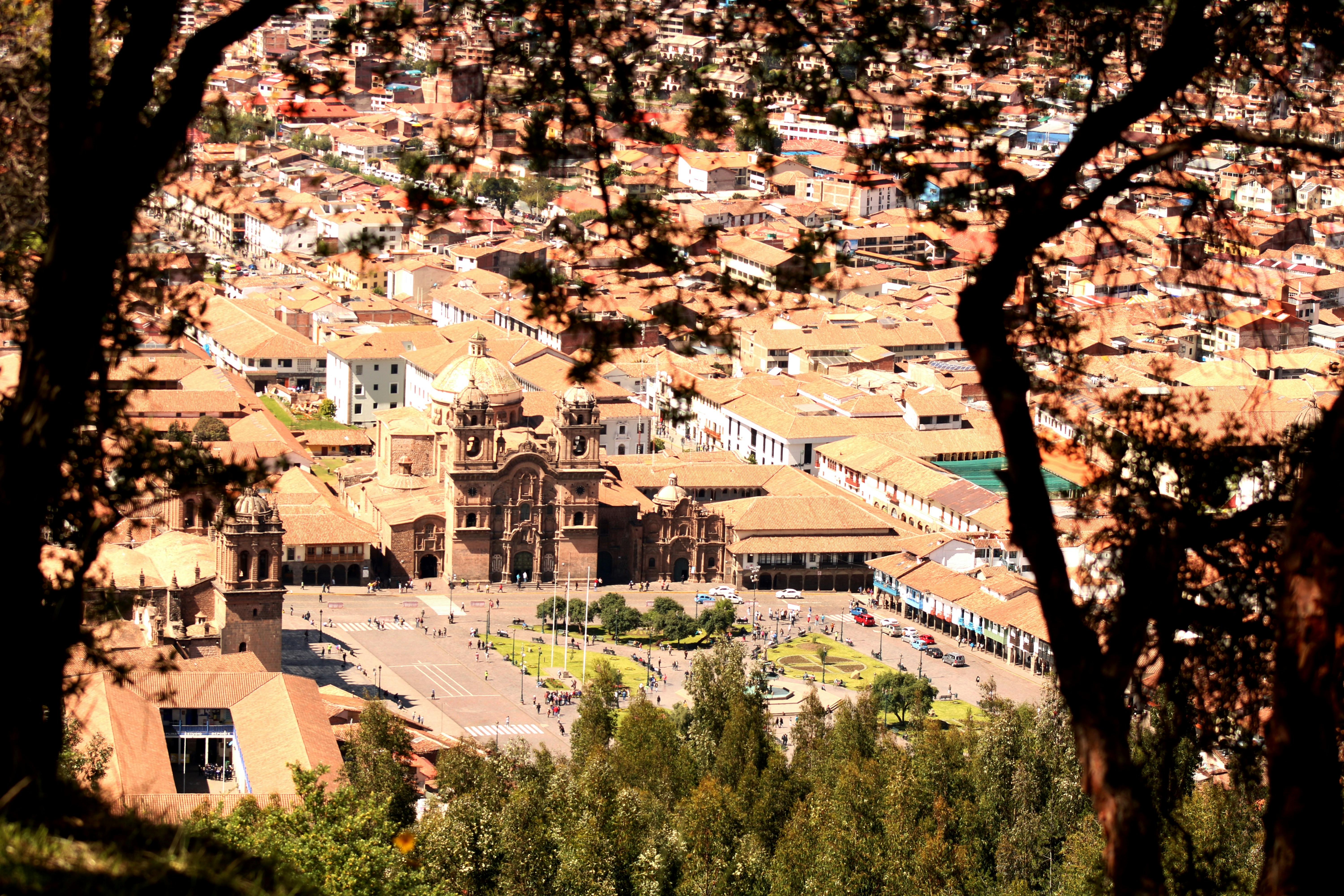 Aerial view of Cusco's historic Plaza de Armas, framed by lush greenery, showcasing colonial architecture and vibrant rooftops. The scene captures the essence of urban life in a historic Peruvian city.