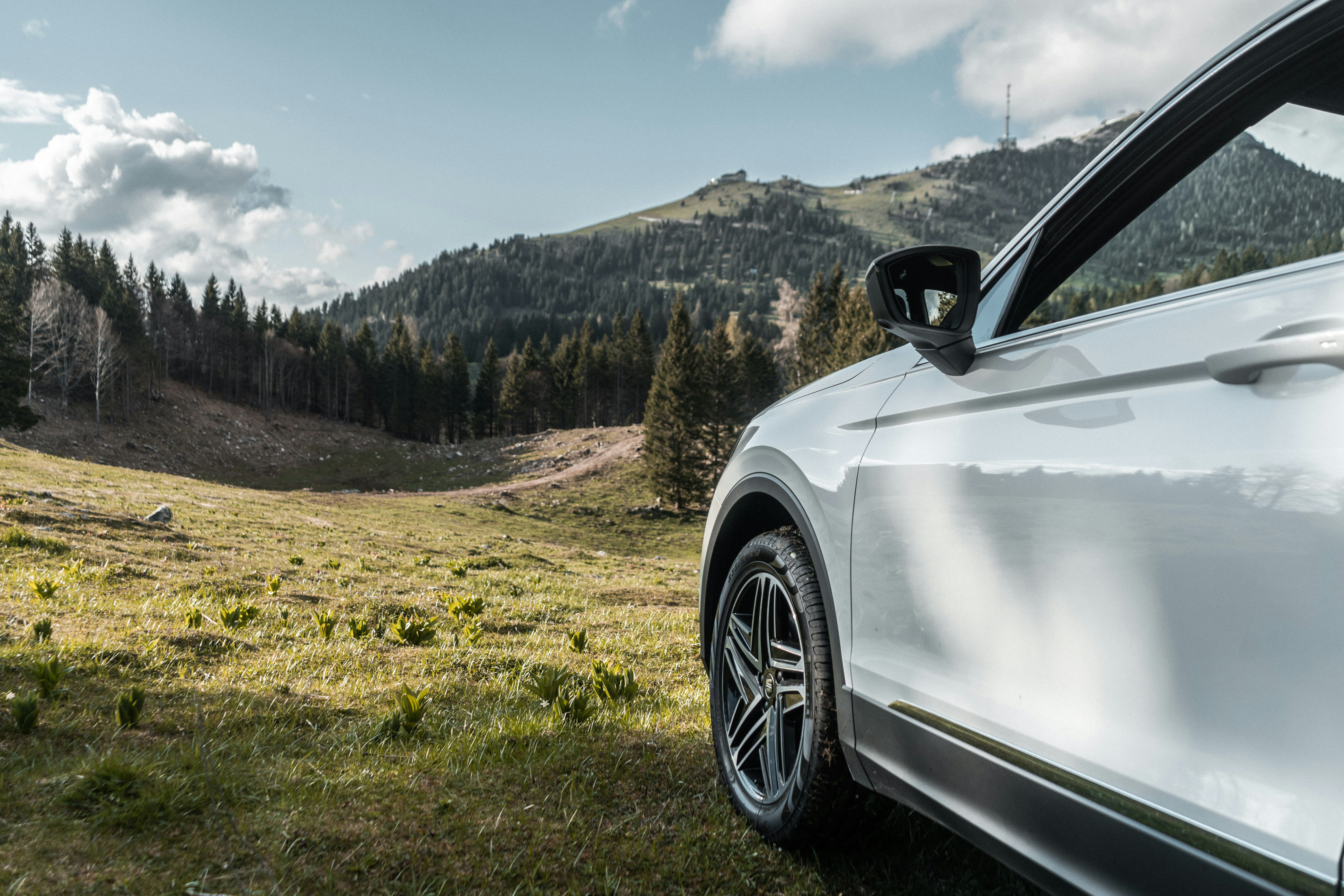 A sleek white car parked on a grassy hillside with a backdrop of lush mountains and scattered clouds. The scene captures the essence of exploration and freedom.