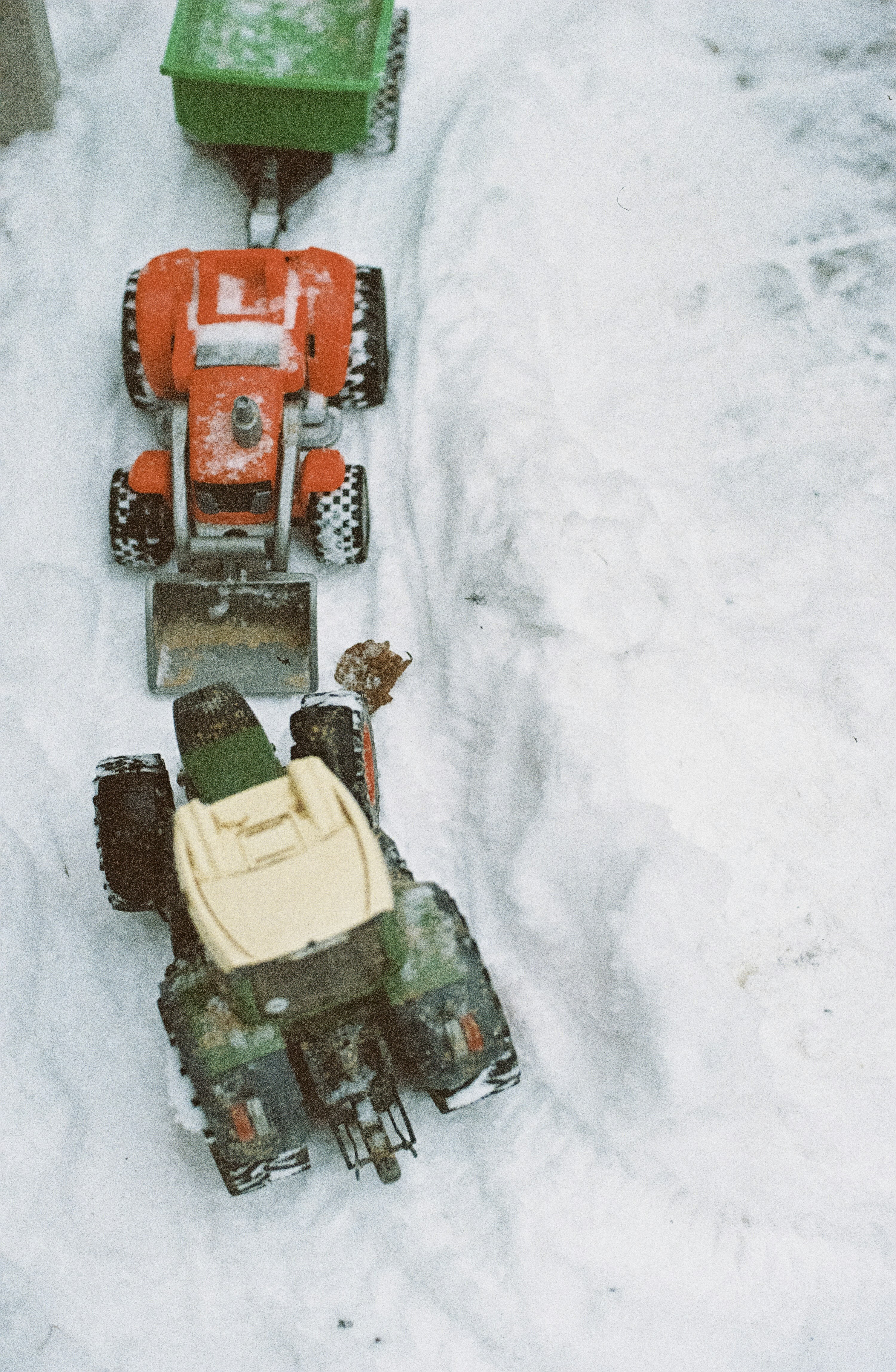Two toy tractors navigating a snowy landscape, with one equipped with a snow plow. The scene captures the essence of winter playtime.