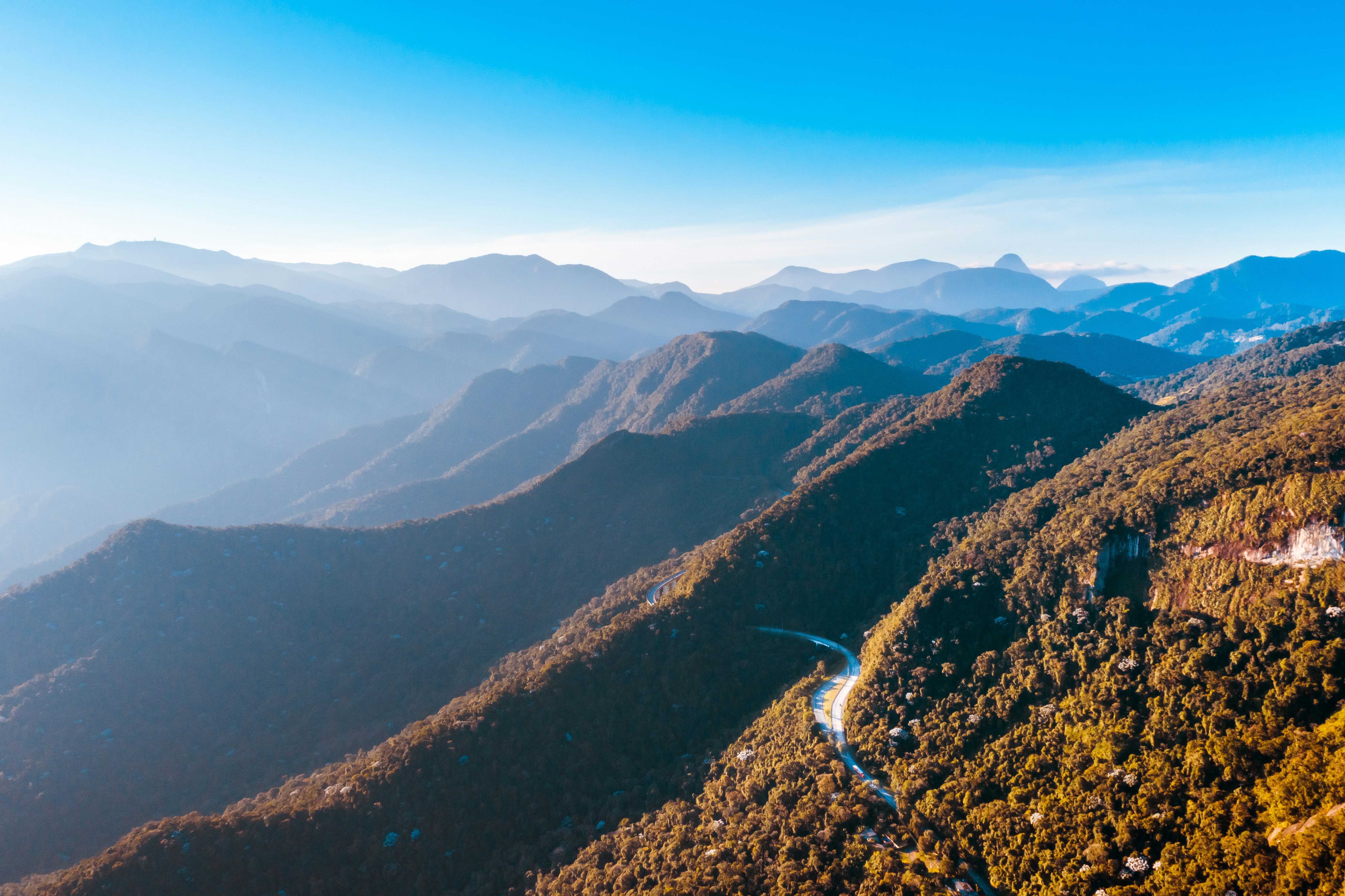 Aerial view of sunlit mountain ridges with a winding road cutting through lush foliage under a clear blue sky.
