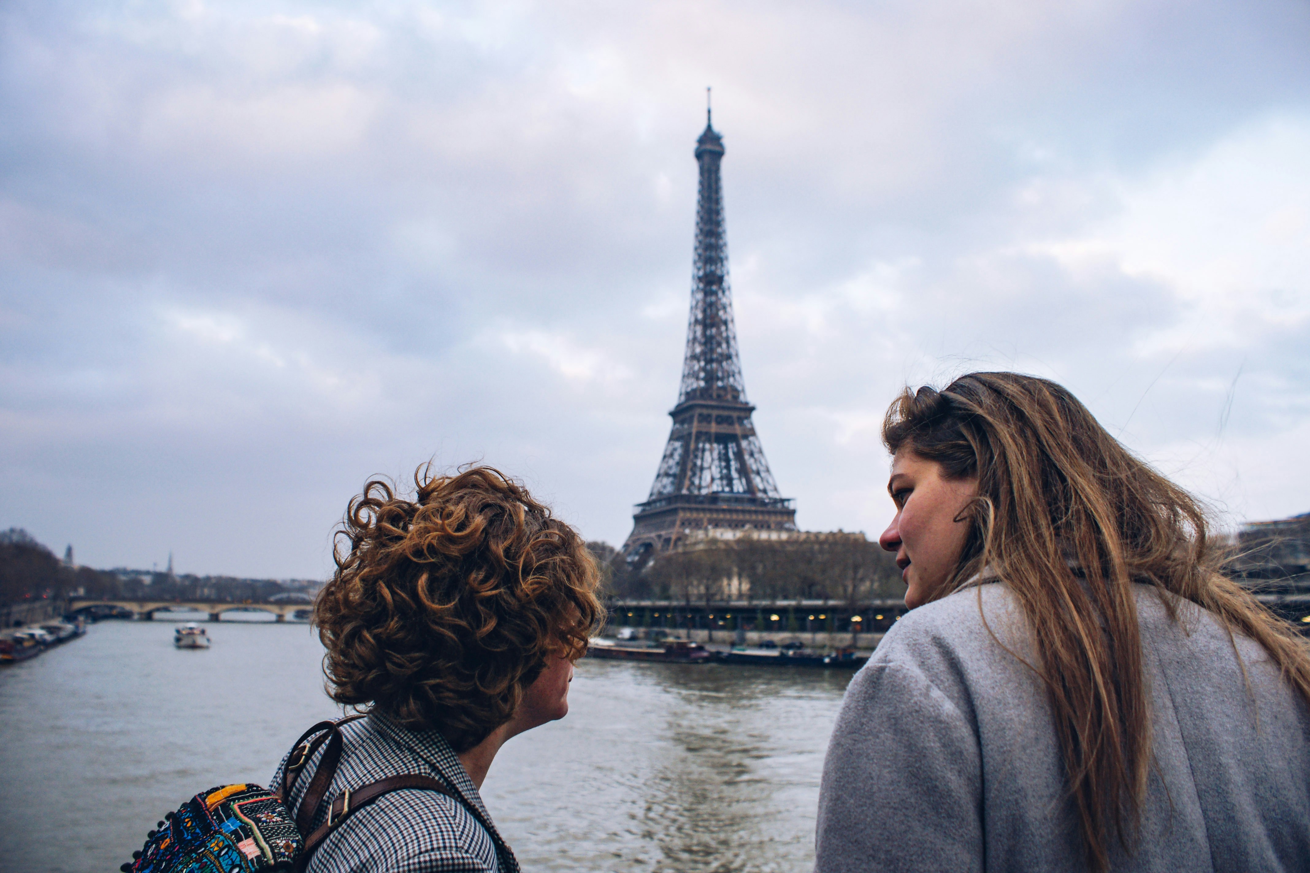 Two travelers stand at the Seine riverbank, gazing toward the Eiffel Tower under a cloudy sky.