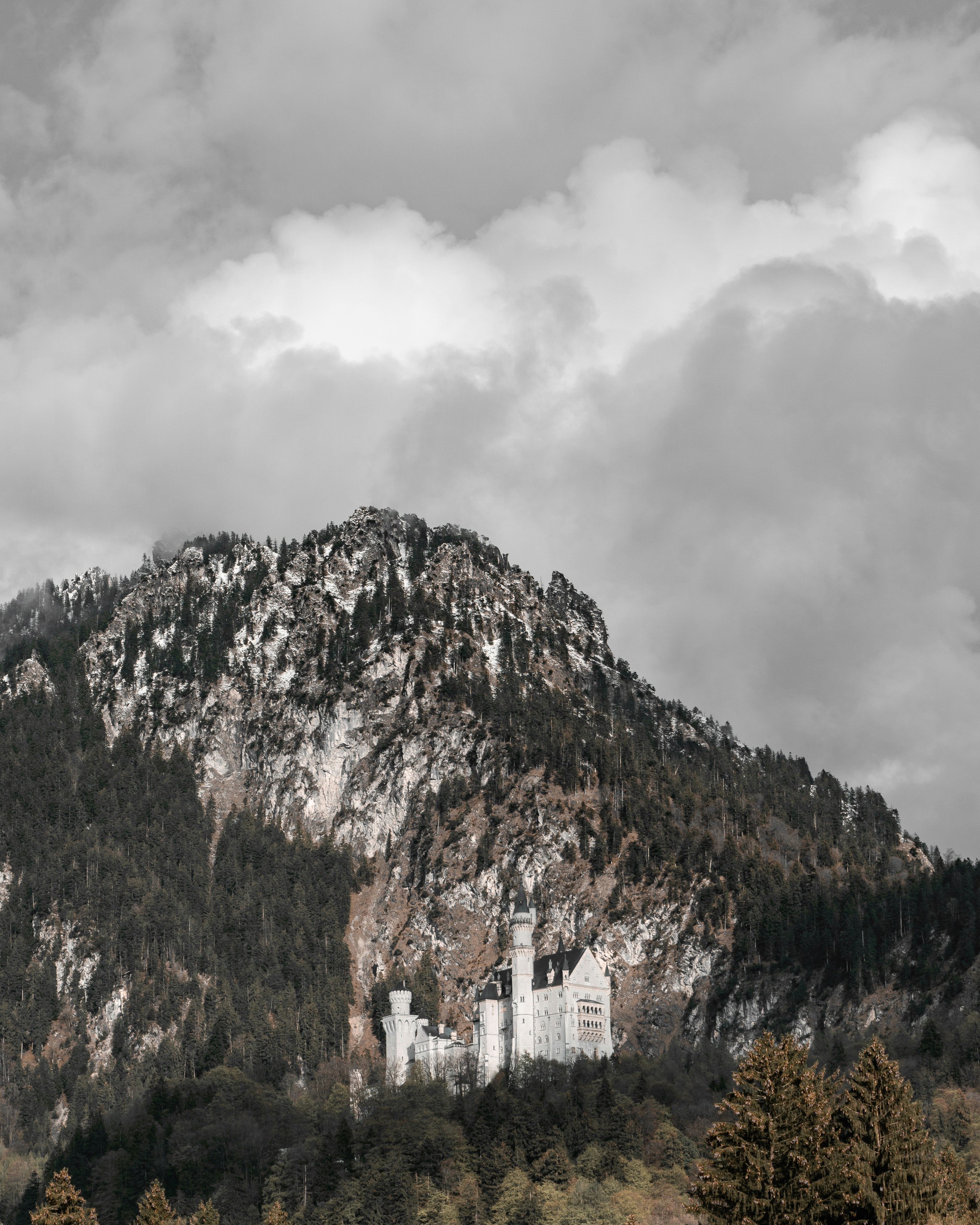 A majestic castle nestled against a rugged mountain backdrop, surrounded by lush greenery and dramatic clouds.