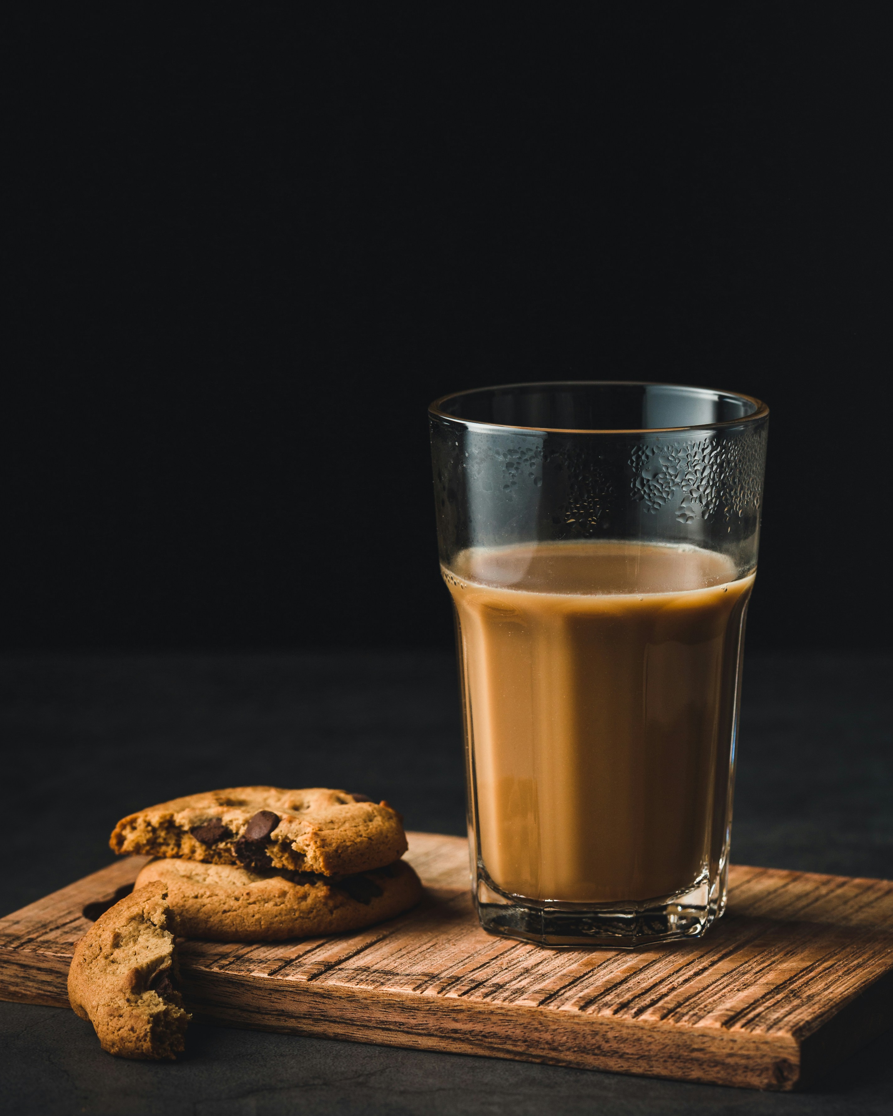 Glass of coffee beside freshly baked chocolate chip cookies on a wooden board against a dark background.