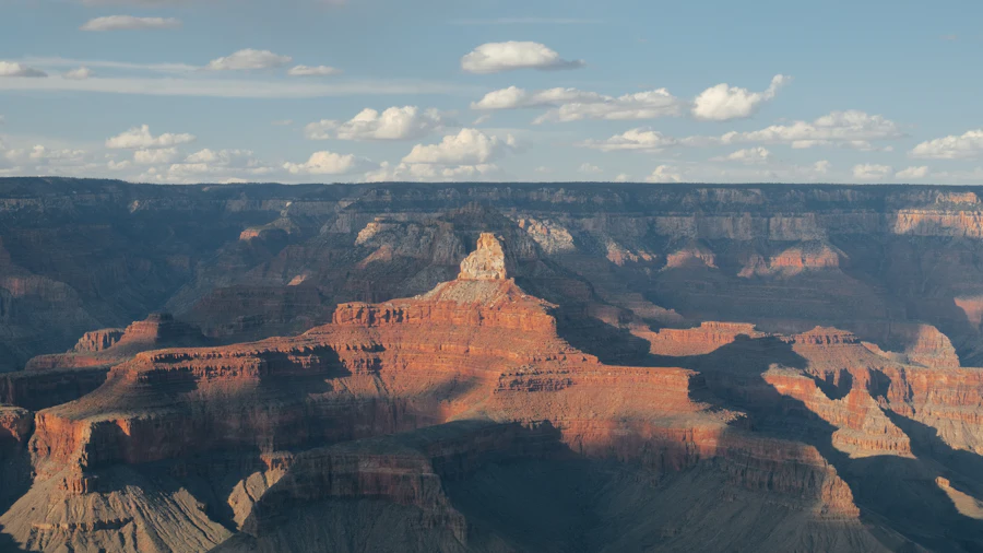 A sunlit red butte rises above the layered cliffs of the Grand Canyon under a blue sky with scattered clouds.