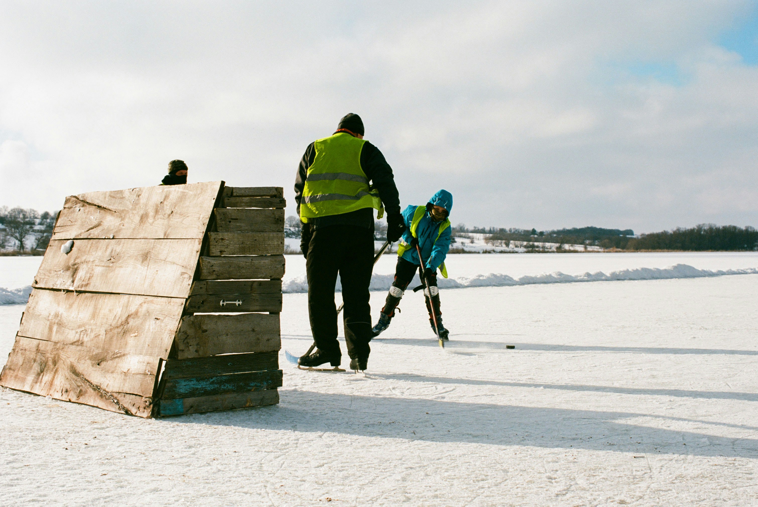 Two figures in high-visibility vests stand by a weathered wooden ramp on a snow-covered field, a child with skis beside an adult. The scene captures a winter coaching moment against a bright, expansive landscape.