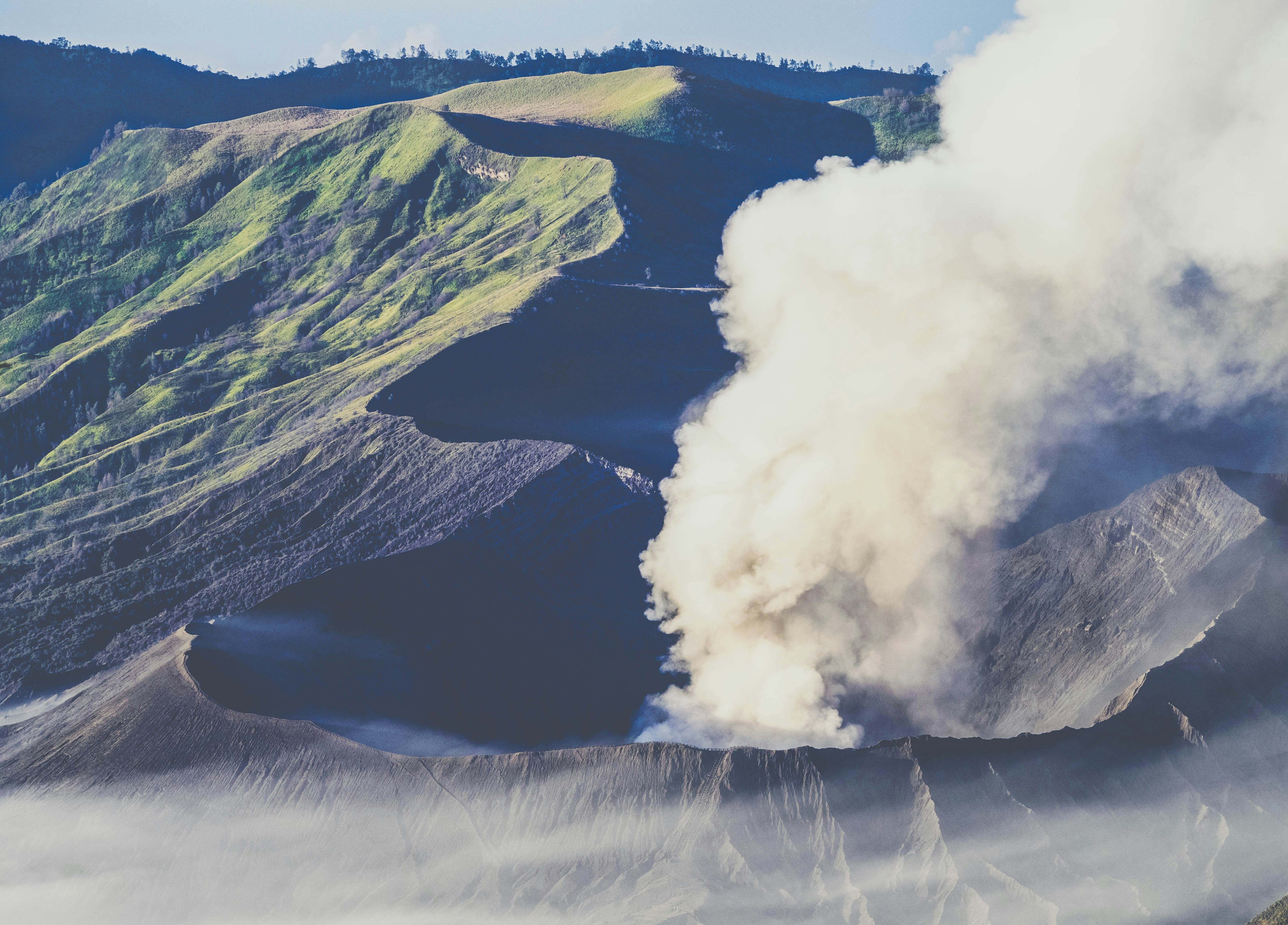 Active volcano emitting a plume of white smoke against rugged green hills under a clear sky.