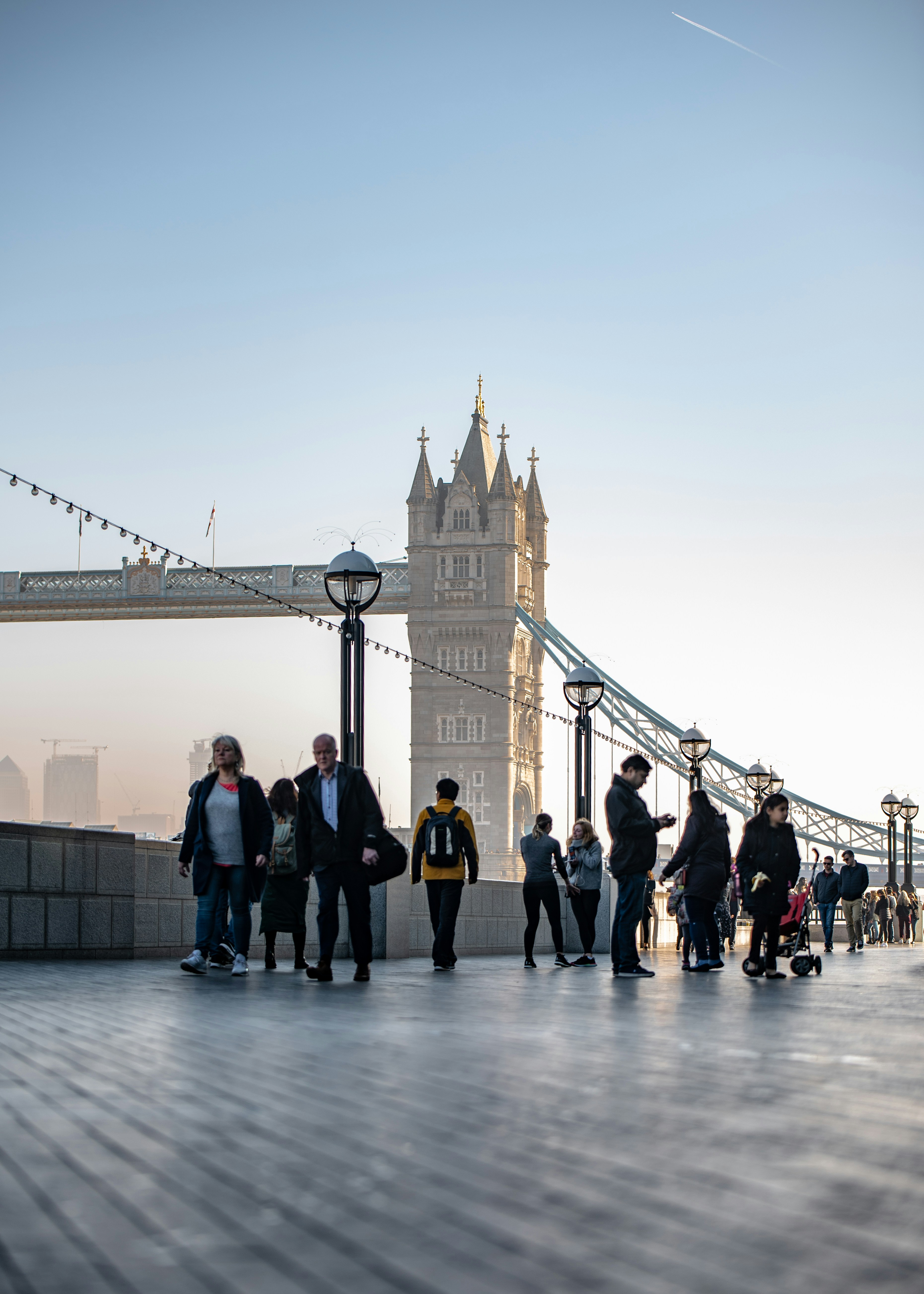 Crowds stroll along the walkway beneath Tower Bridge, with the iconic structure standing tall against a serene sky. 