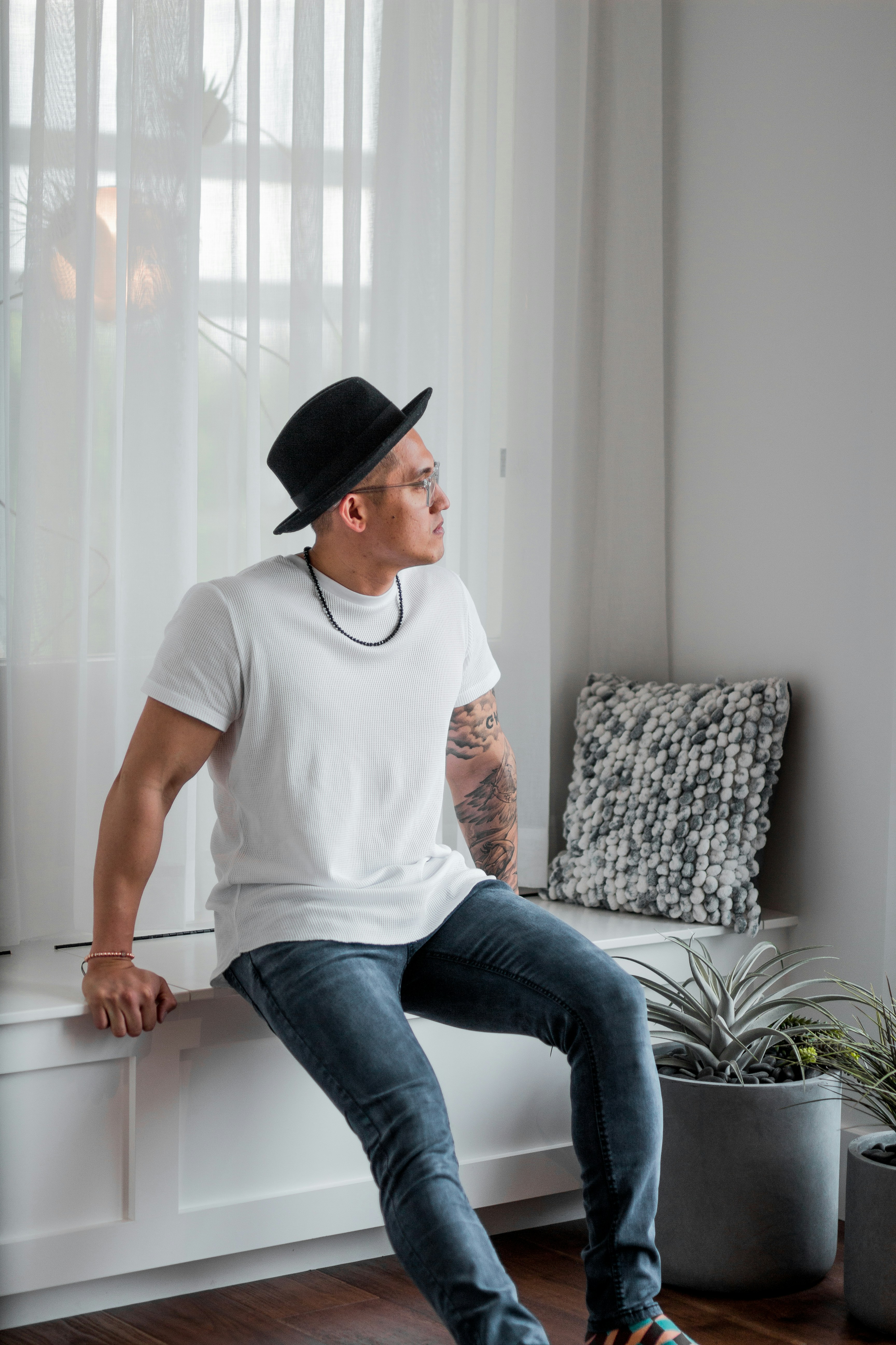 A man in a white t-shirt and hat sits thoughtfully by a window, surrounded by elegant decor and greenery.