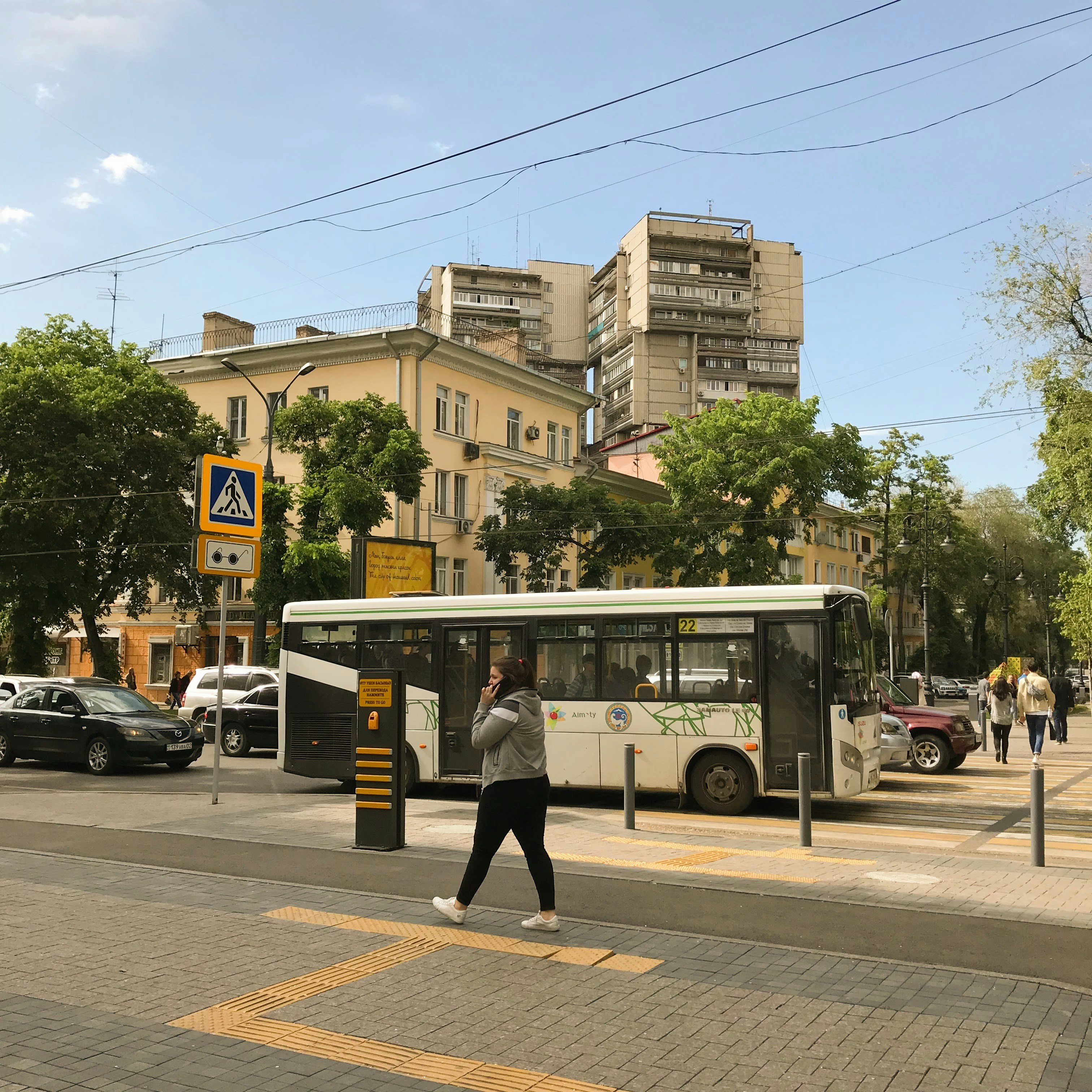 A city bus stops at a bustling intersection, surrounded by trees and modern architecture, with pedestrians navigating the urban landscape.