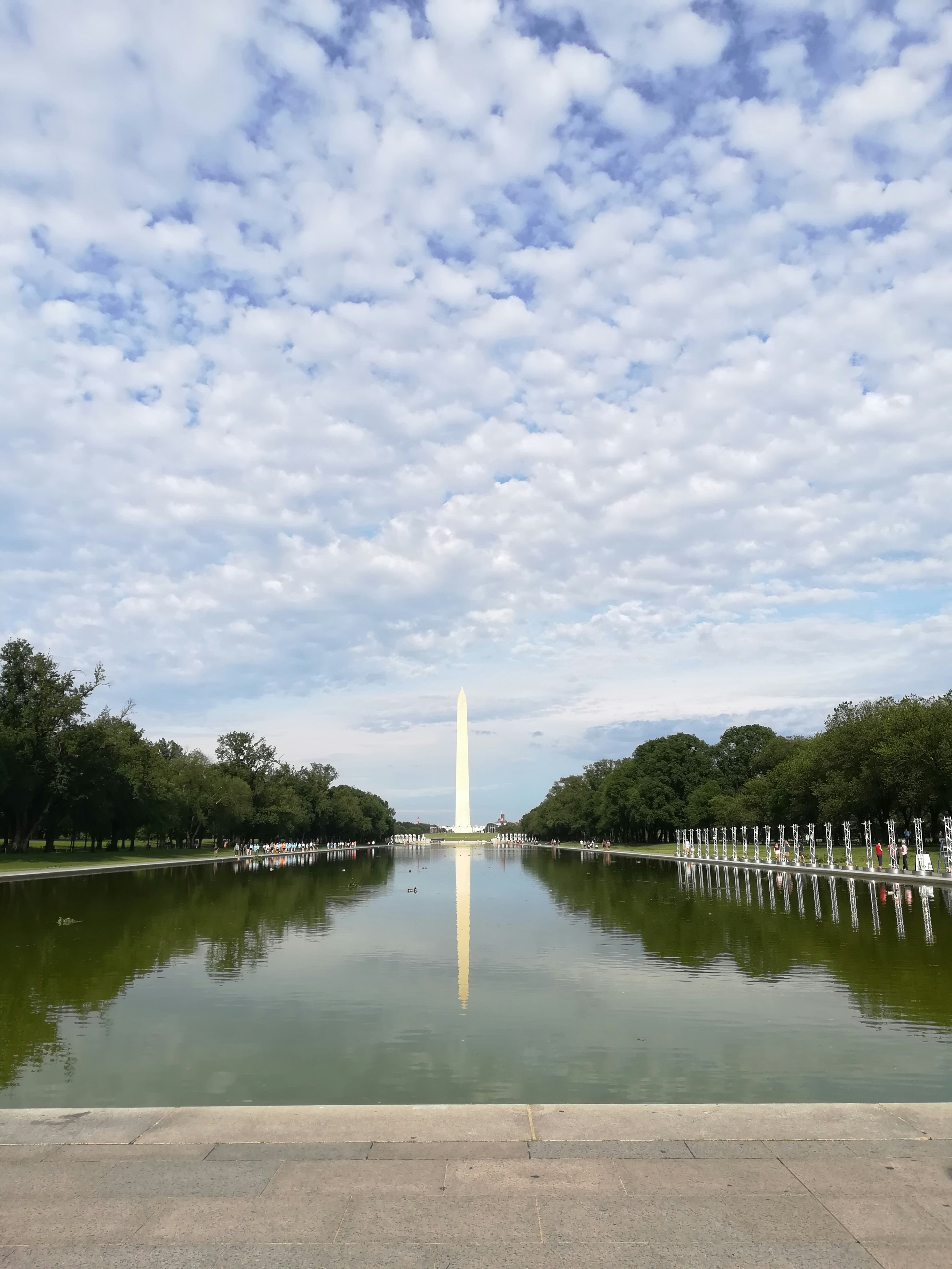 The Washington Monument reflects in the calm waters of the reflecting pool, framed by lush greenery and a dramatic sky.