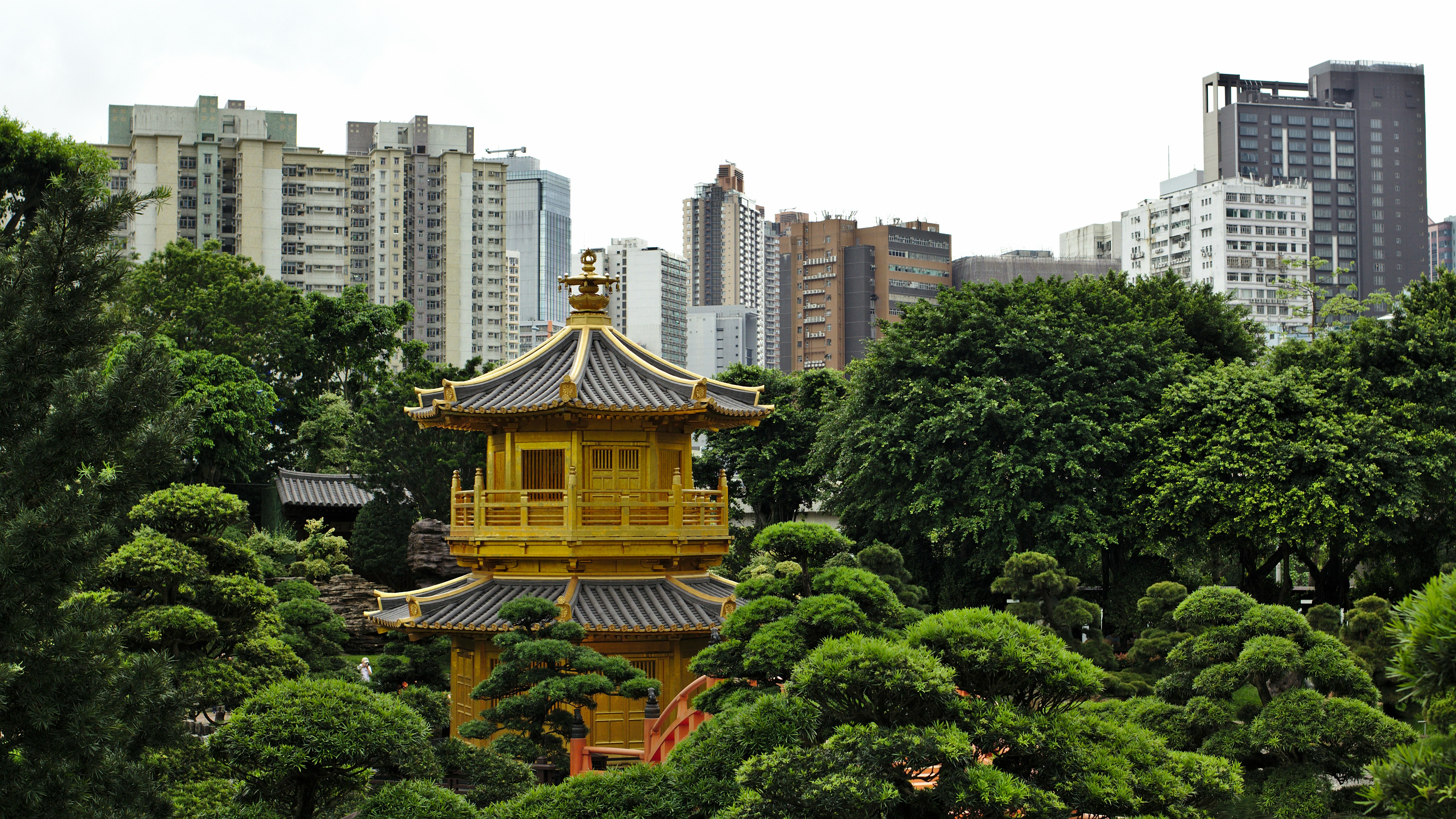 Golden pavilion nestled among lush greenery with a backdrop of towering skyscrapers. The scene illustrates the contrast between traditional architecture and modern city life.